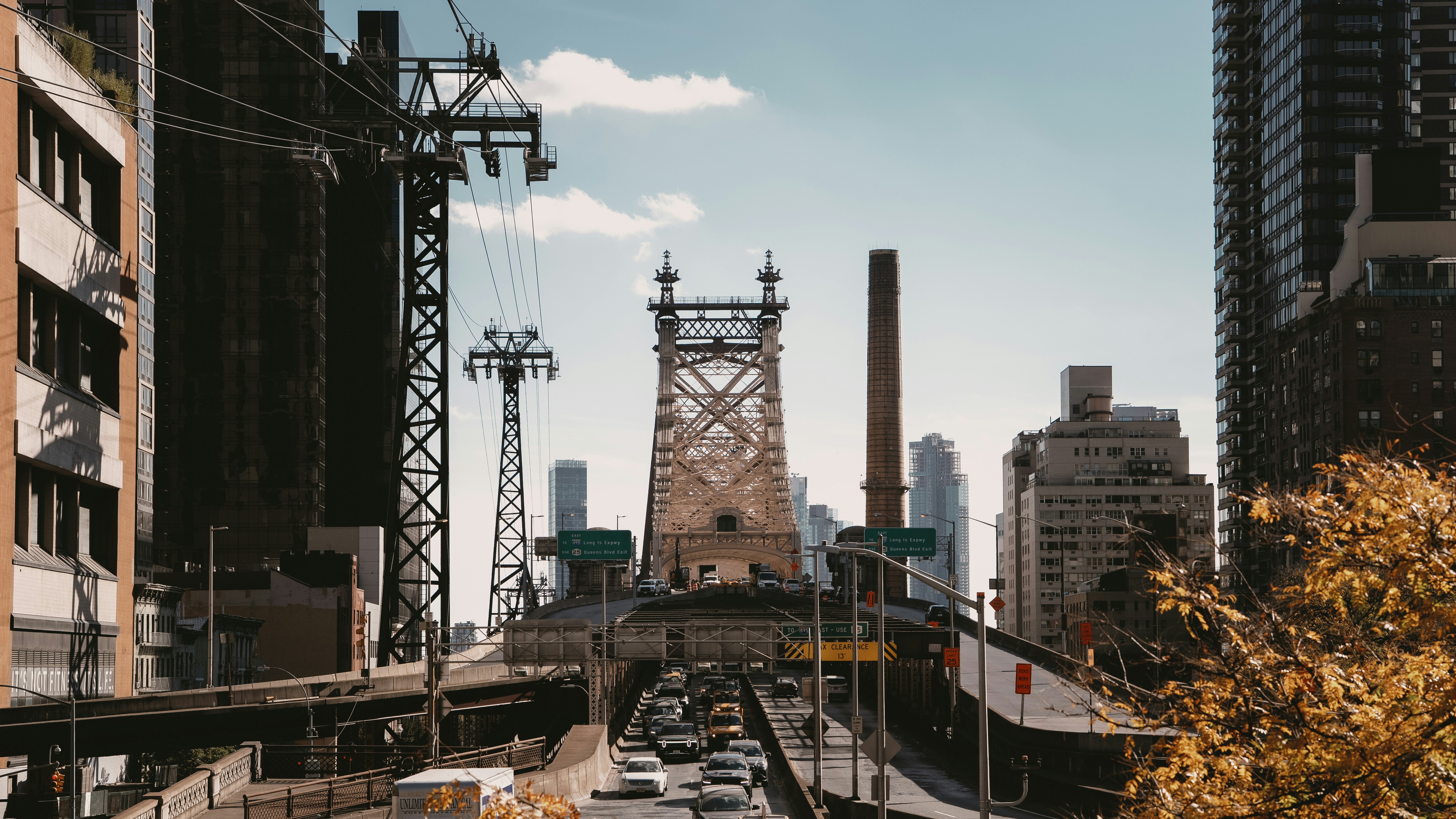 The iconic Queensboro Bridge towers over a bustling cityscape, framed by modern skyscrapers and vibrant autumn foliage.