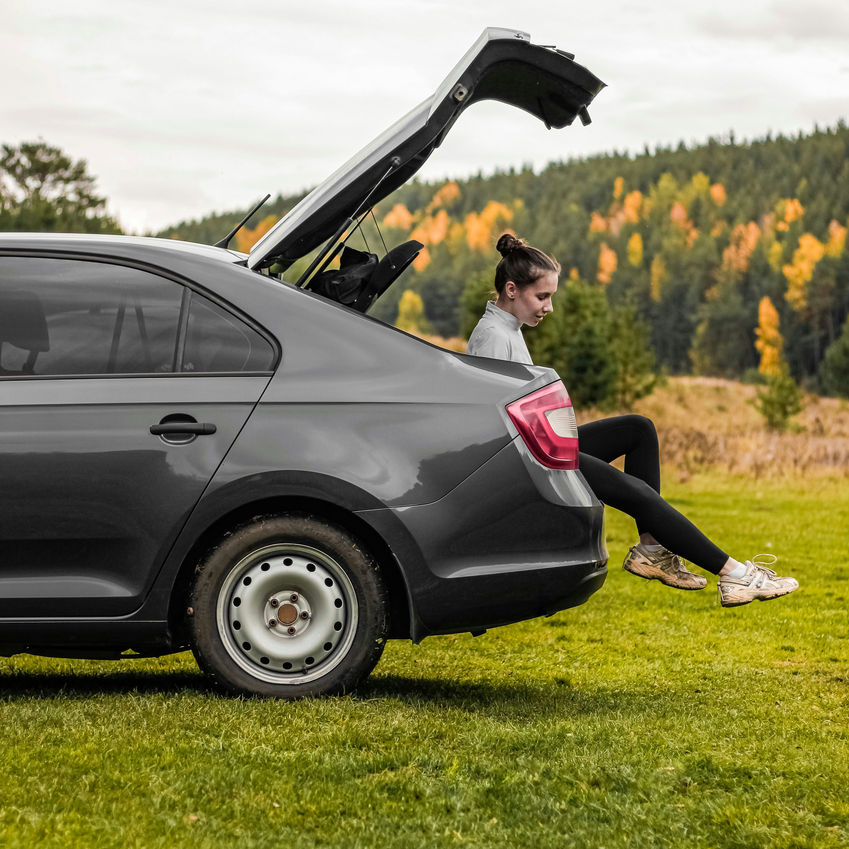 A young woman sits on the edge of a car's trunk, enjoying the tranquility of a lush landscape with autumn foliage in the background.