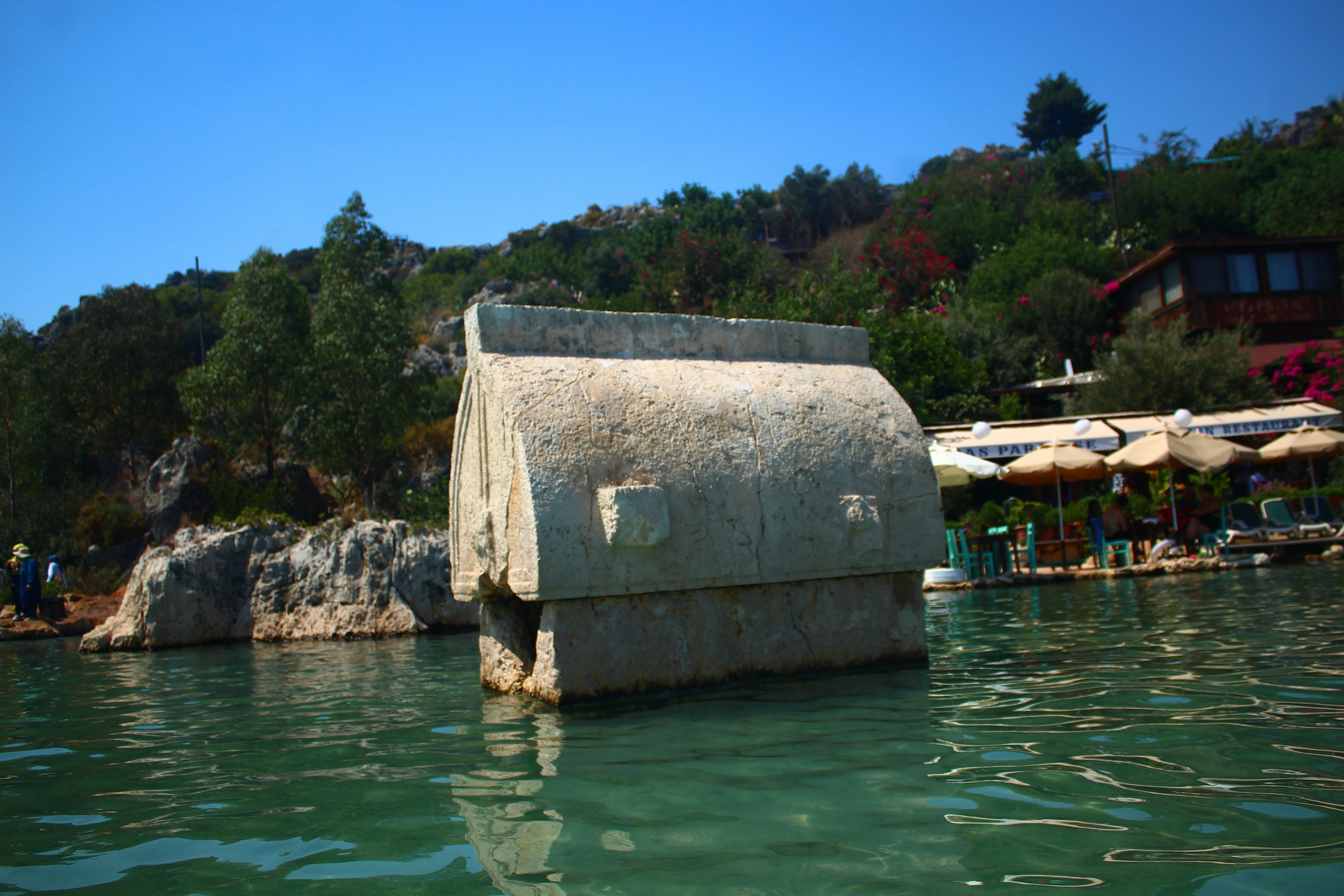 Weathered stone structure partially submerged in clear turquoise water, surrounded by lush greenery and a rustic café setting.