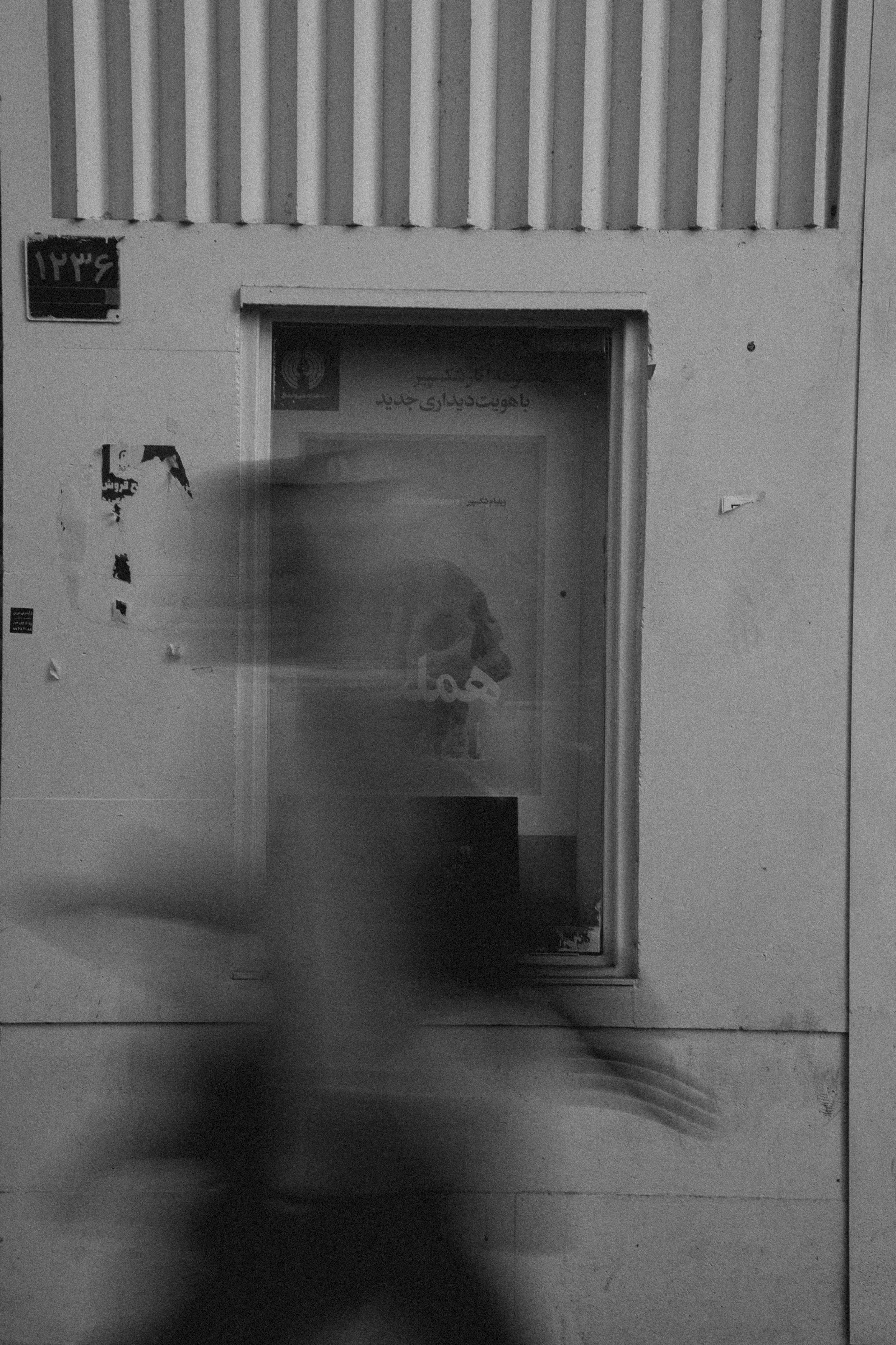 A blurred figure passing by a storefront, capturing the essence of movement in a city setting. The monochrome tones enhance the atmosphere of fleeting moments.