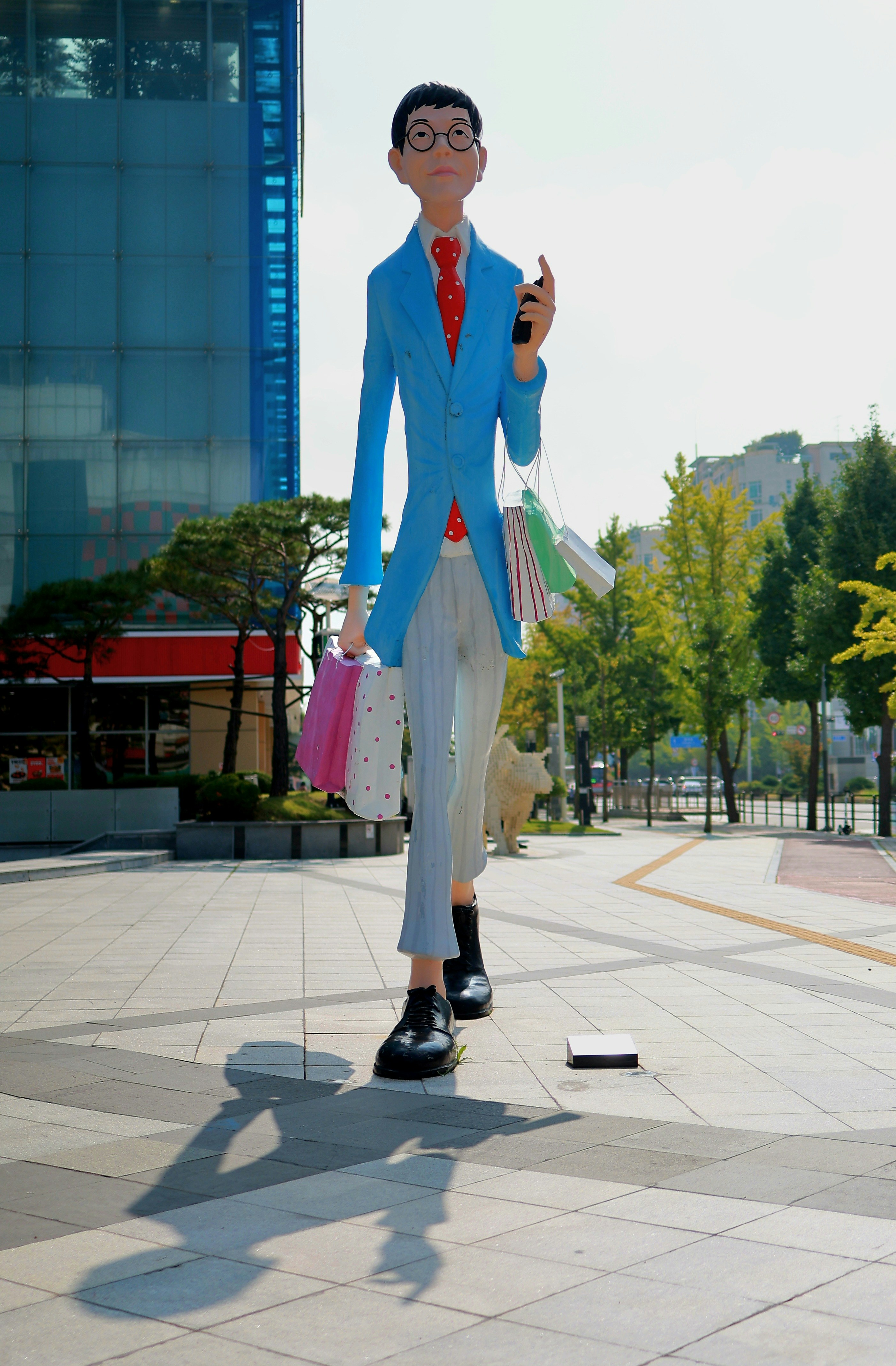 Tall cartoon figure in a blue suit holding shopping bags, walking through an urban plaza with trees and a modern building in the background.