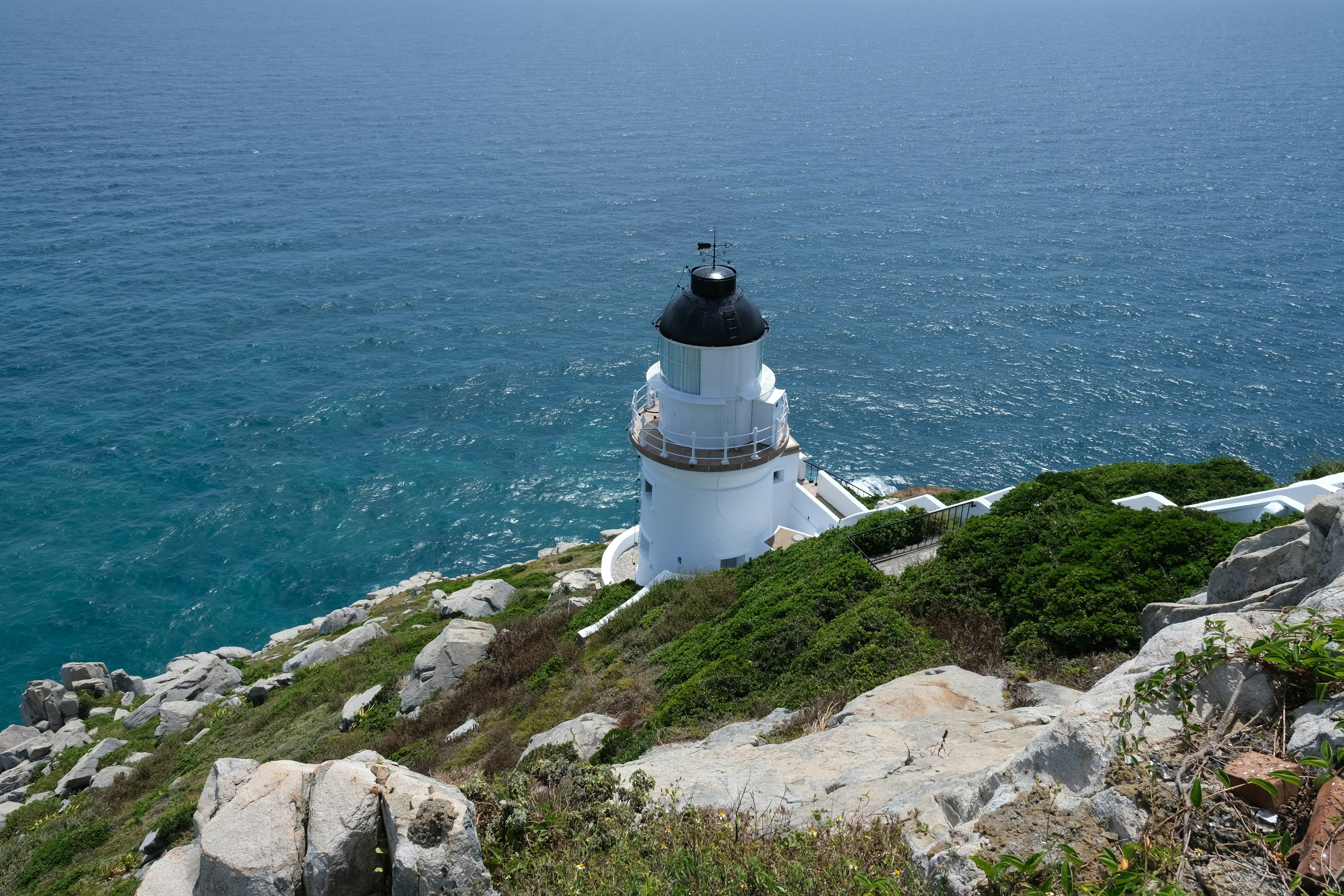 A lighthouse perched on rocky cliffs overlooking the azure sea, surrounded by lush greenery. The structure stands as a sentinel against the vast ocean.