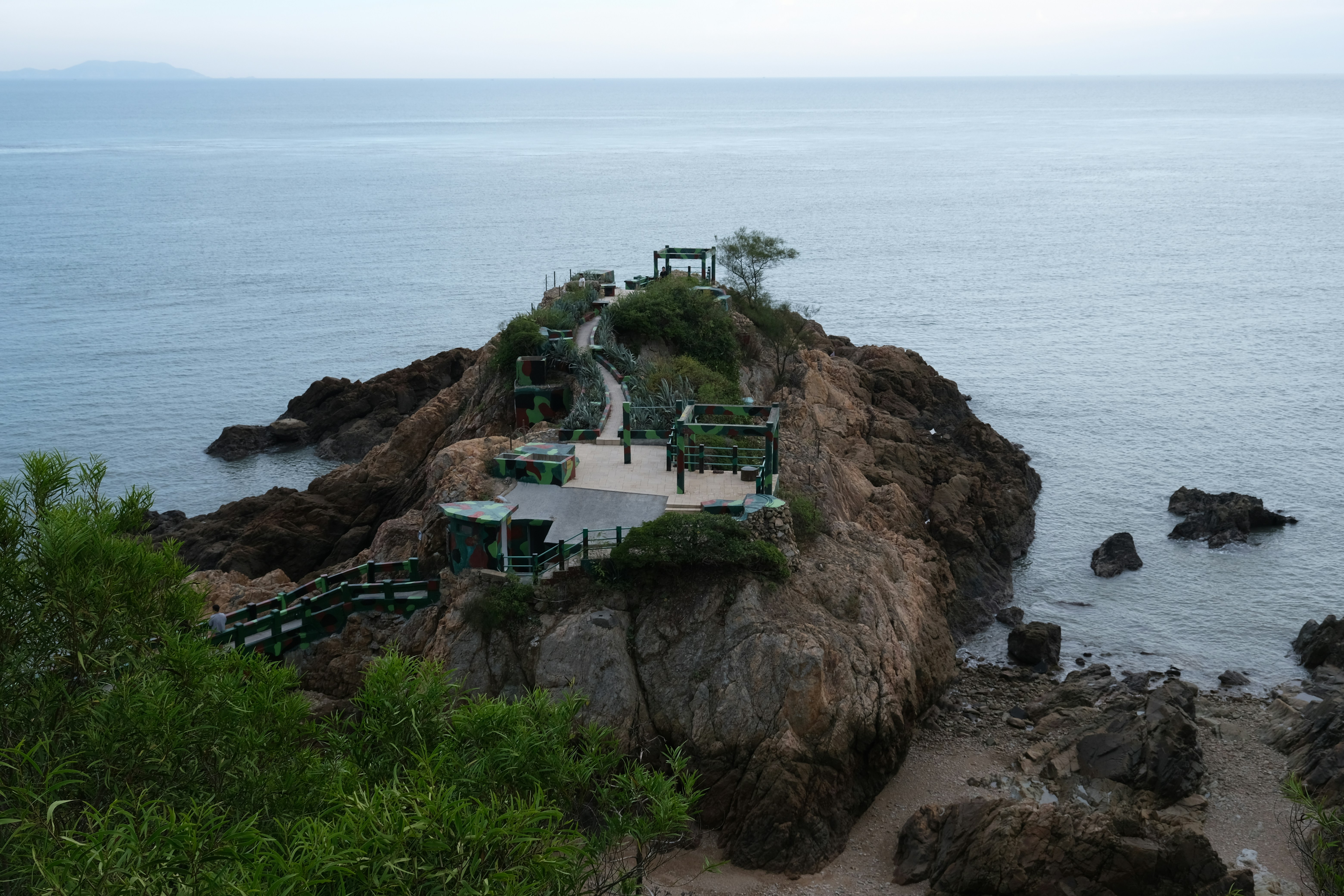 A winding pathway leads to a viewing platform atop a rocky outcrop, surrounded by calm waters and lush greenery.