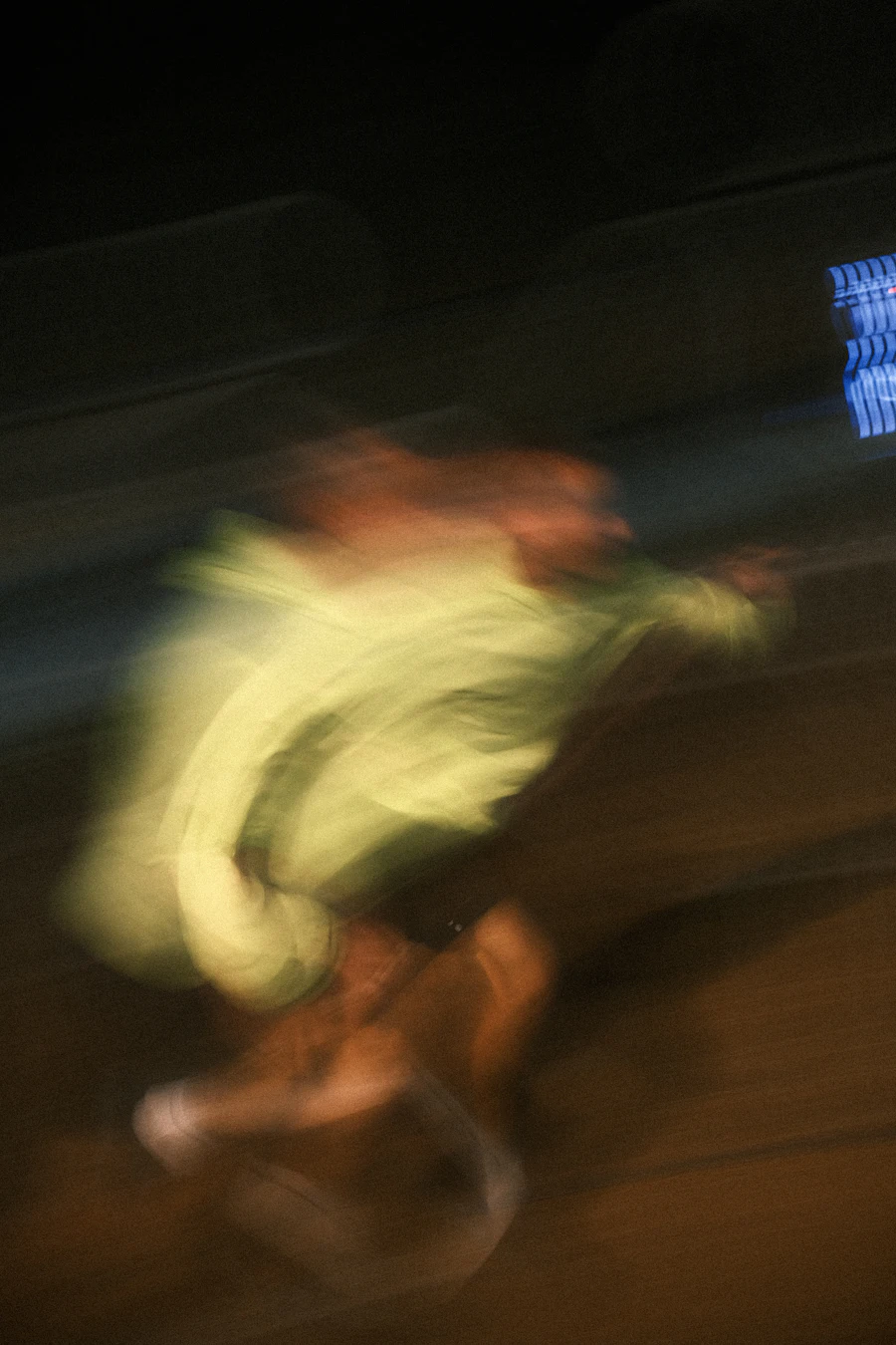 A cyclist in a bright jacket streaks through the dark with heavy motion blur, creating a sense of speed and movement. Blue lights glow on the right edge of the frame.