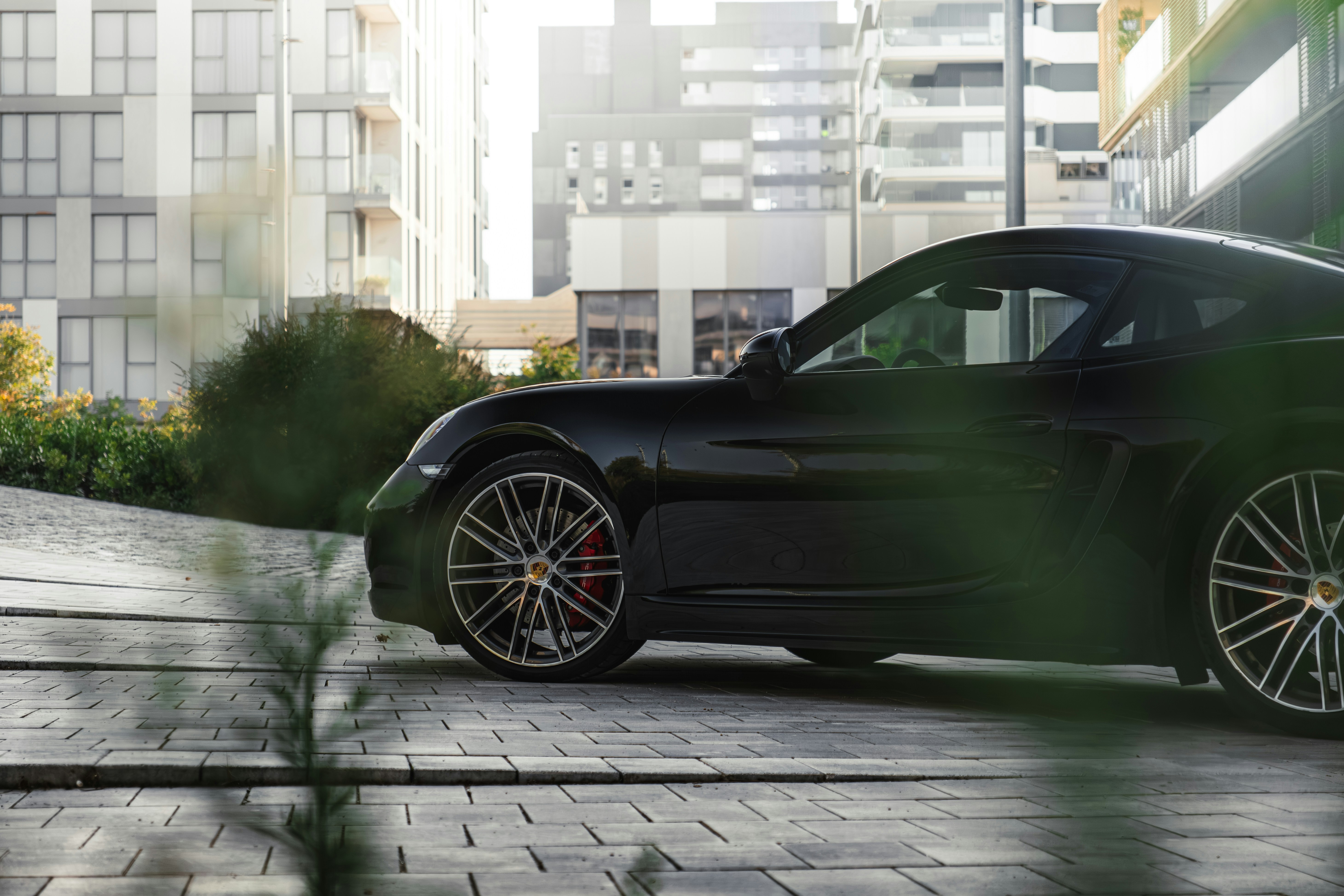 A sleek black sports car parked on a contemporary urban street, framed by modern buildings and greenery. The composition highlights the car's design against a backdrop of architectural lines.