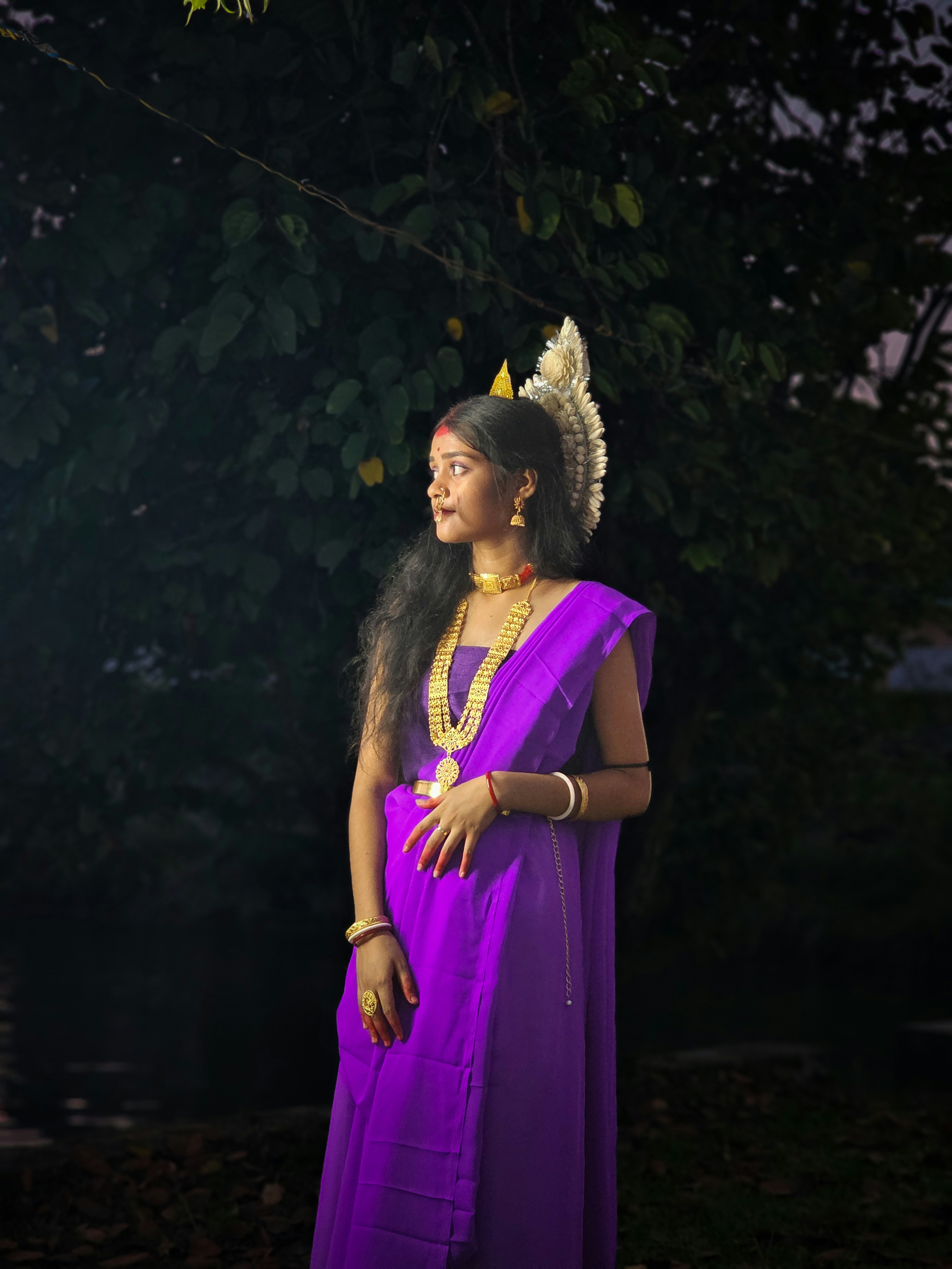 Young woman adorned in vibrant purple attire and traditional jewelry, standing gracefully against a lush backdrop. The soft lighting highlights her features beautifully.