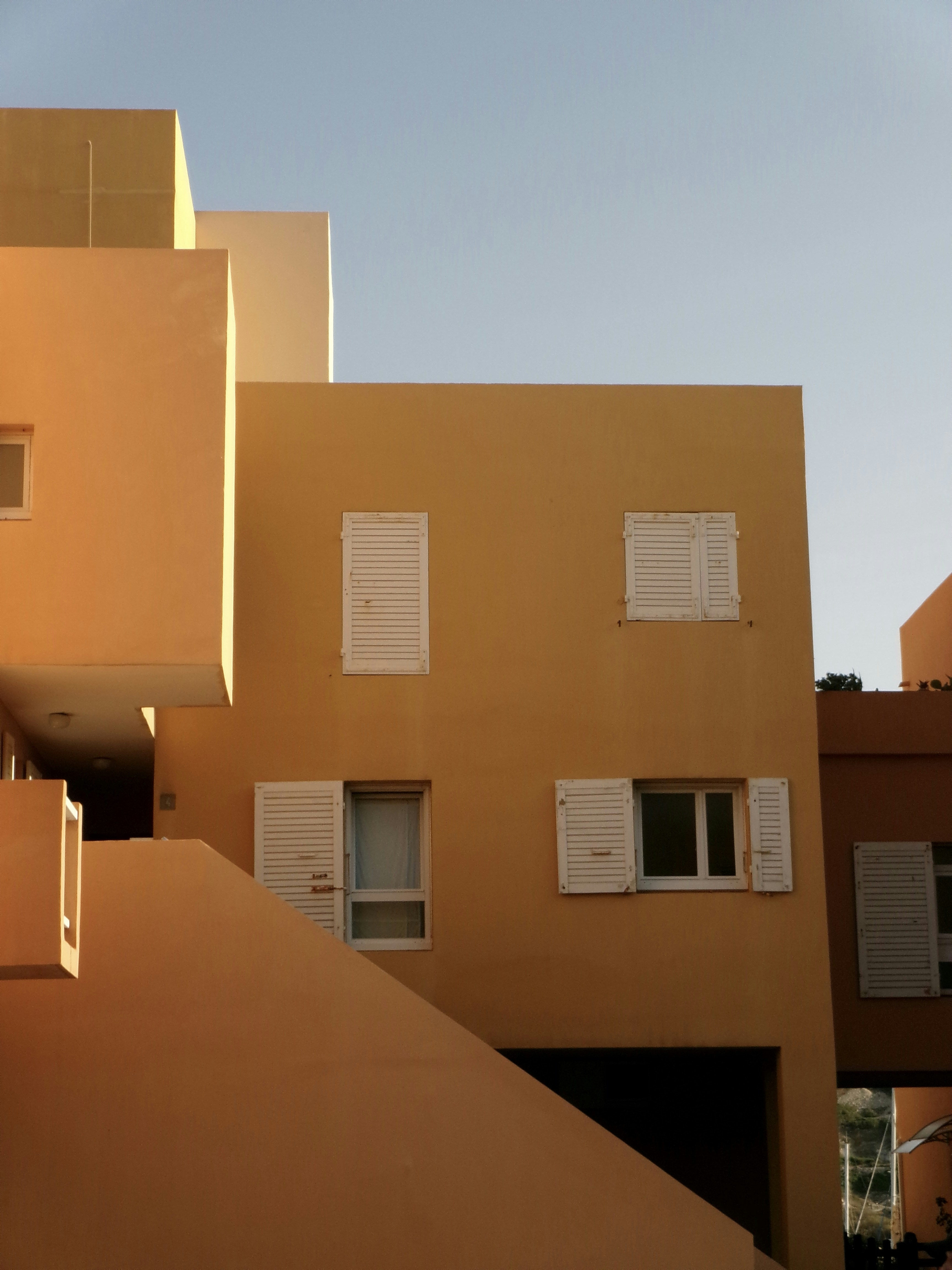 Vibrant yellow building facade with white shutters and geometric lines, bathed in soft sunlight. Architectural design highlights the interplay of light and shadow.