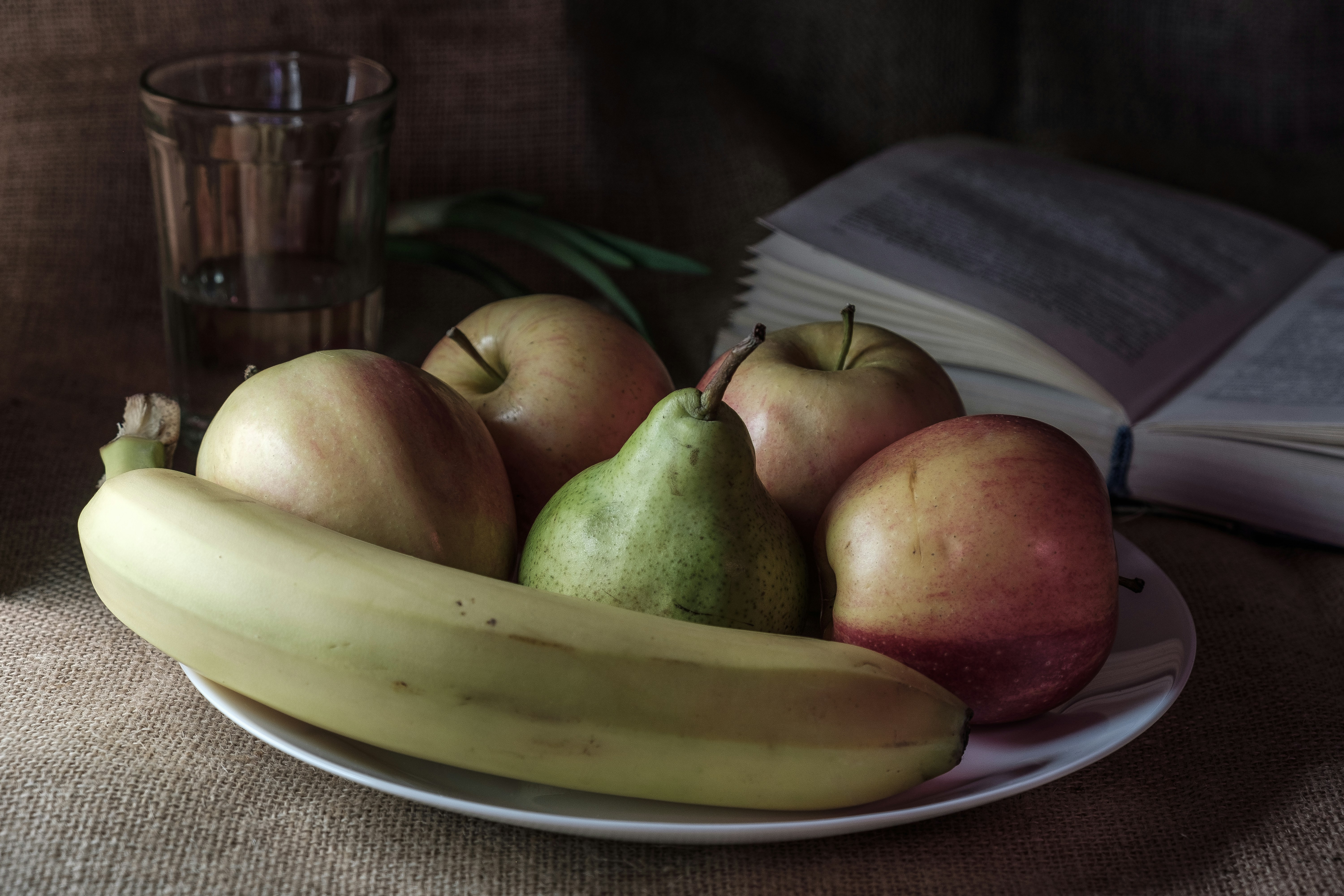 still life with apples pear and banana in the background is a glass with a book | Still life of fruit with a glass and book.