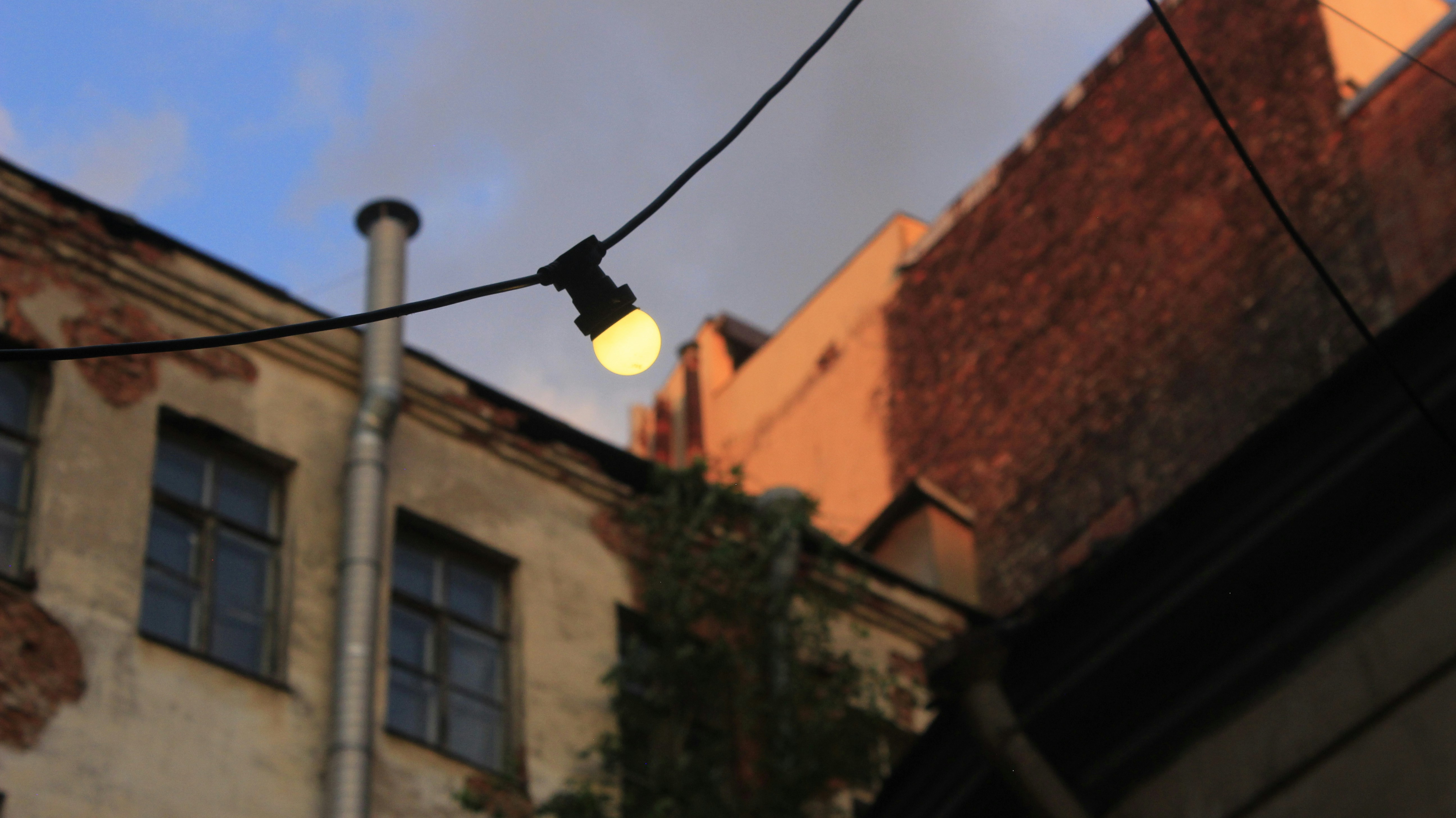 Glowing bulb hanging from a wire against a backdrop of weathered buildings and a dusky sky.