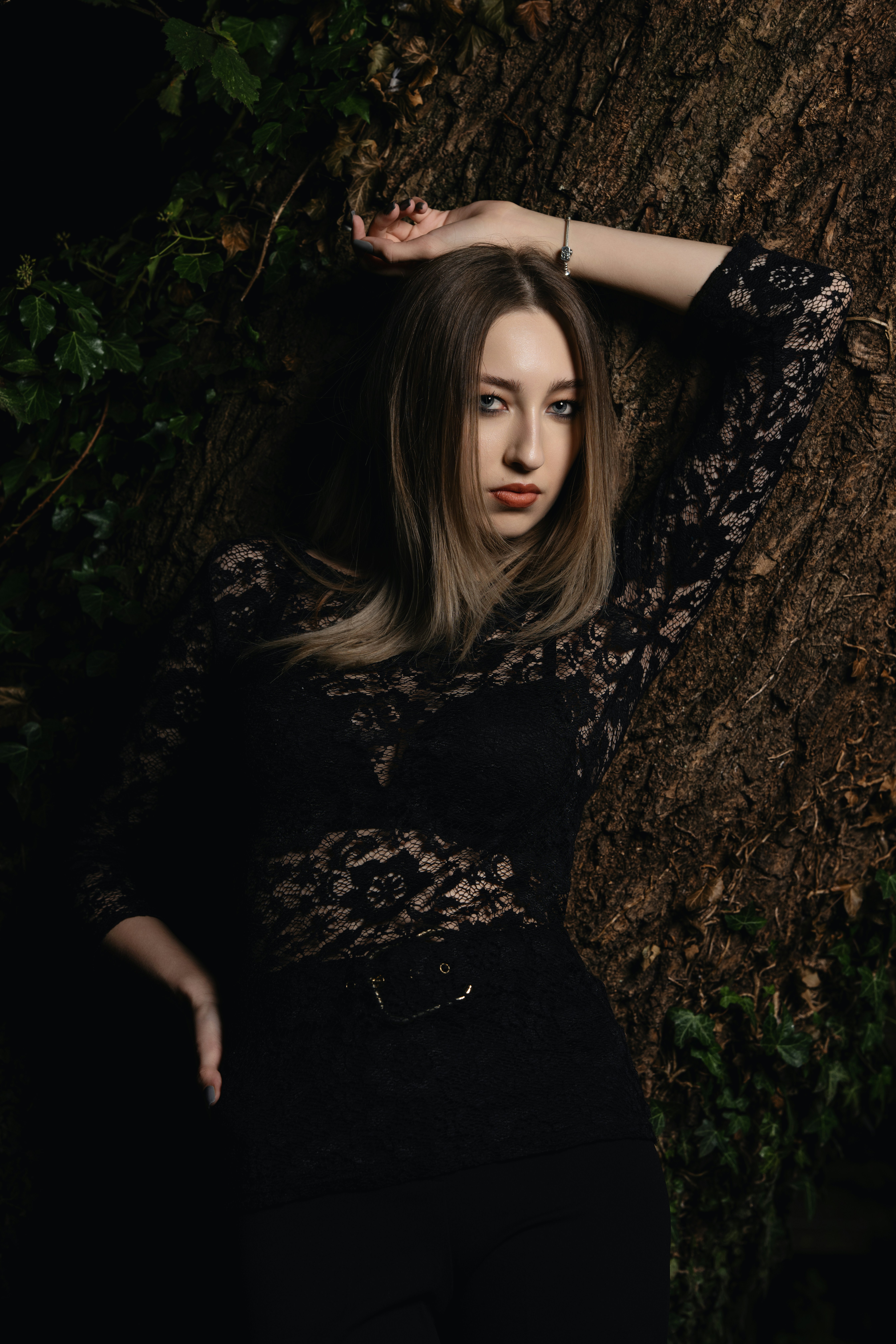 Model poses against a textured tree trunk, draped in delicate black lace, with soft, dramatic lighting enhancing her features.