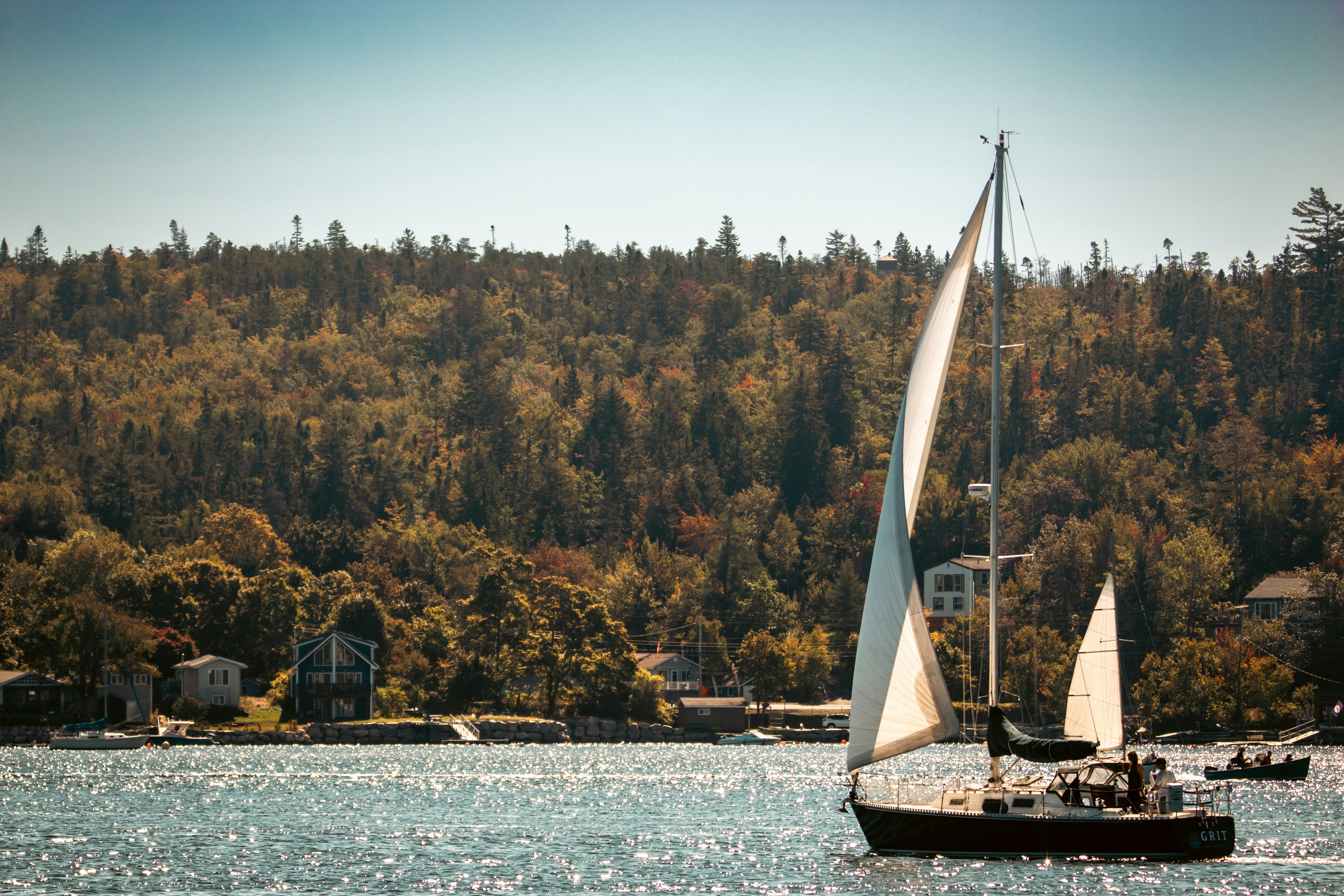 A sailboat glides through sparkling waters, framed by a backdrop of vibrant autumn foliage and quaint lakeside homes.