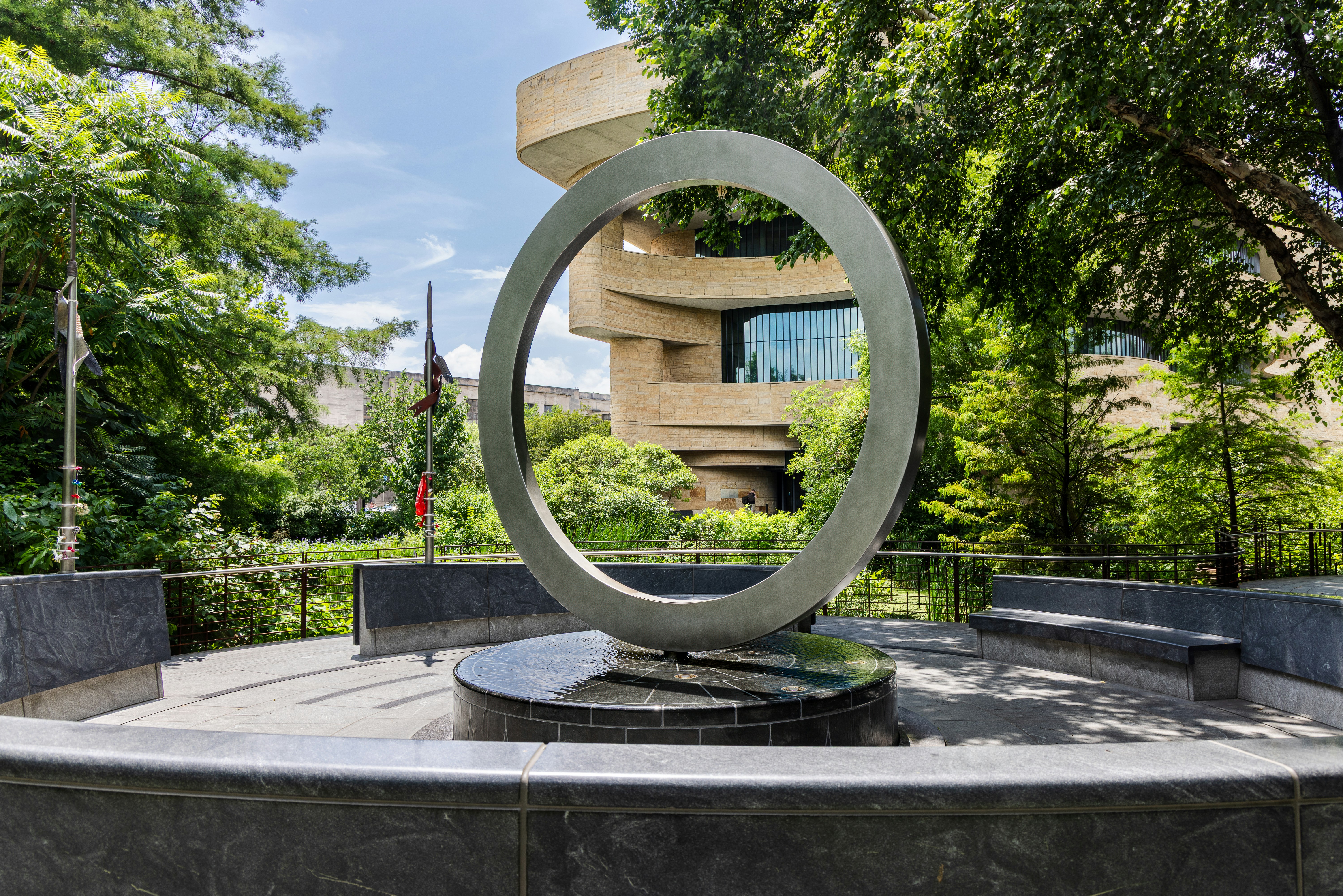 The National Native American Veterans Memorial opened on November 11, 2020, on the grounds of the National Museum of the American Indian in Washington, DC. The memorial was dedicated with a procession and ceremony on the National Mall on November 11, 2022. This tribute to Native heroes recognizes for the first time on a national scale the enduring and distinguished service of Native Americans in every branch of the US military. | Modern sculpture in a park with building background