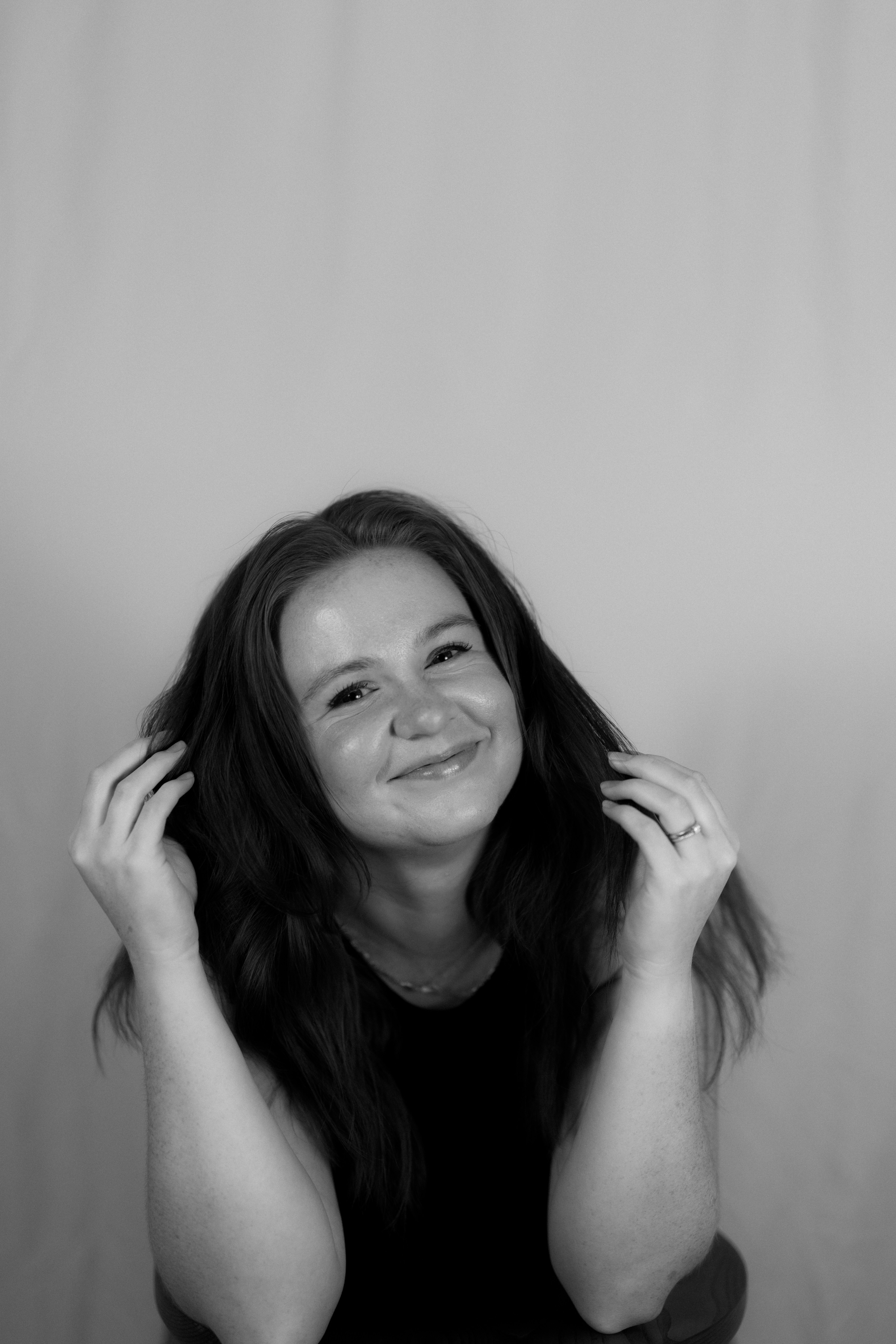 A smiling young woman playfully tousles her hair against a soft gray backdrop.