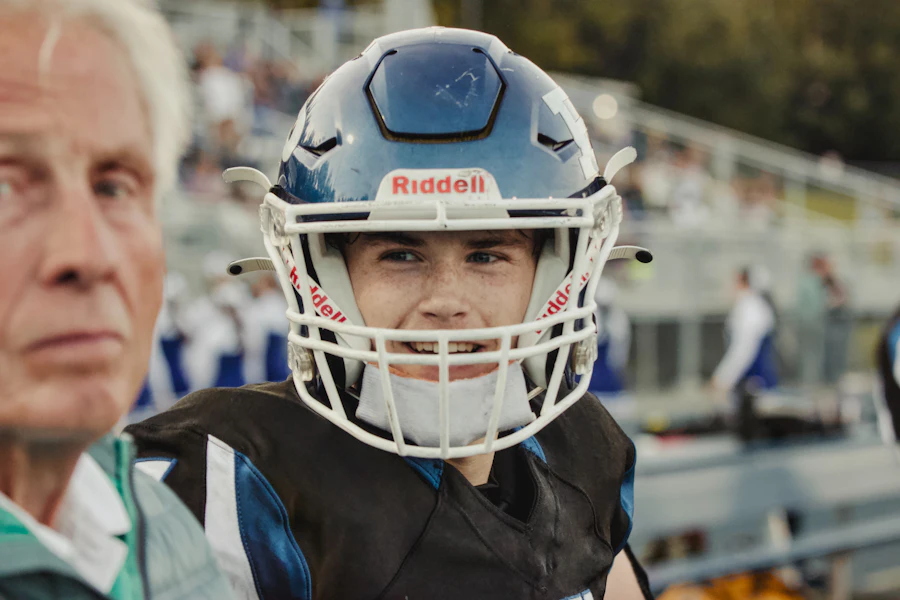 Young football player in helmet smiling on the sideline with a blurred coach in the foreground and bleachers behind.