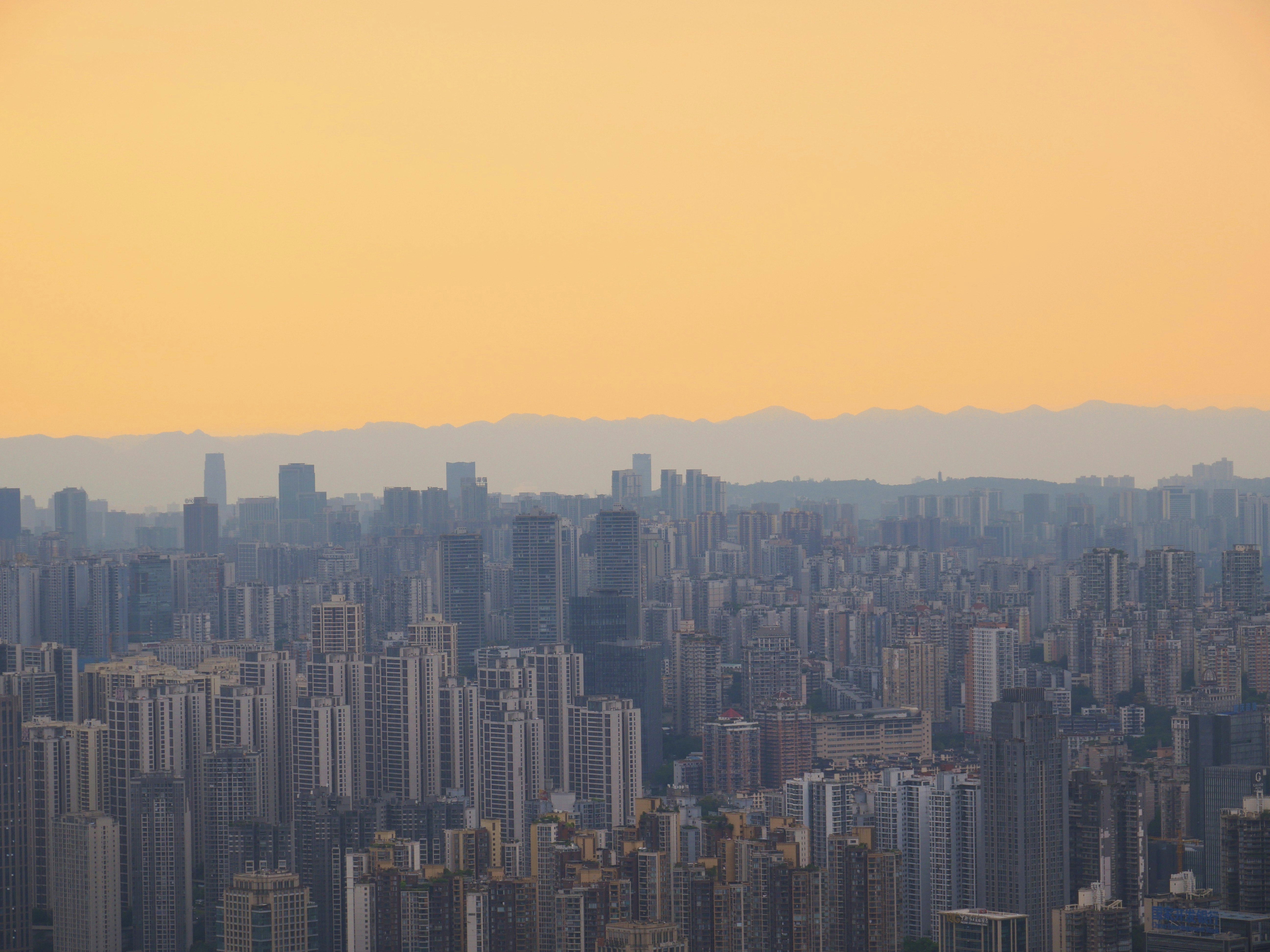 Spectacular sunset over Chongqing's modern skyline in China, captured from a high vantage point. The golden-orange sky creates a dramatic backdrop for the sprawling urban landscape of towering skyscrapers and dense residential buildings, showcasing one of China's major metropolitan areas during the magical golden hour. | City skyline with hazy mountains at sunset