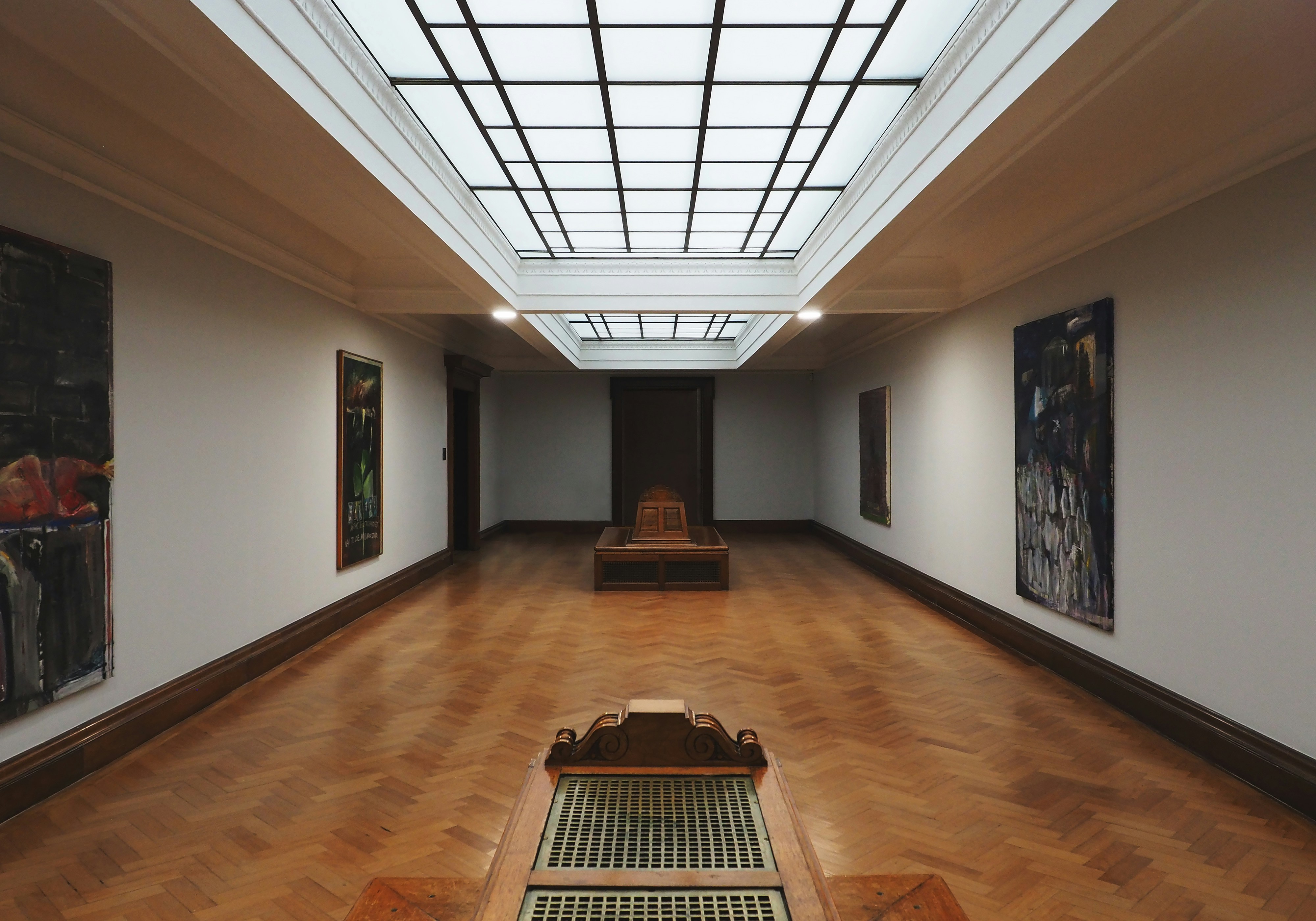 Minimalist art gallery featuring wooden flooring and illuminated skylight, framed by abstract paintings on the walls.
