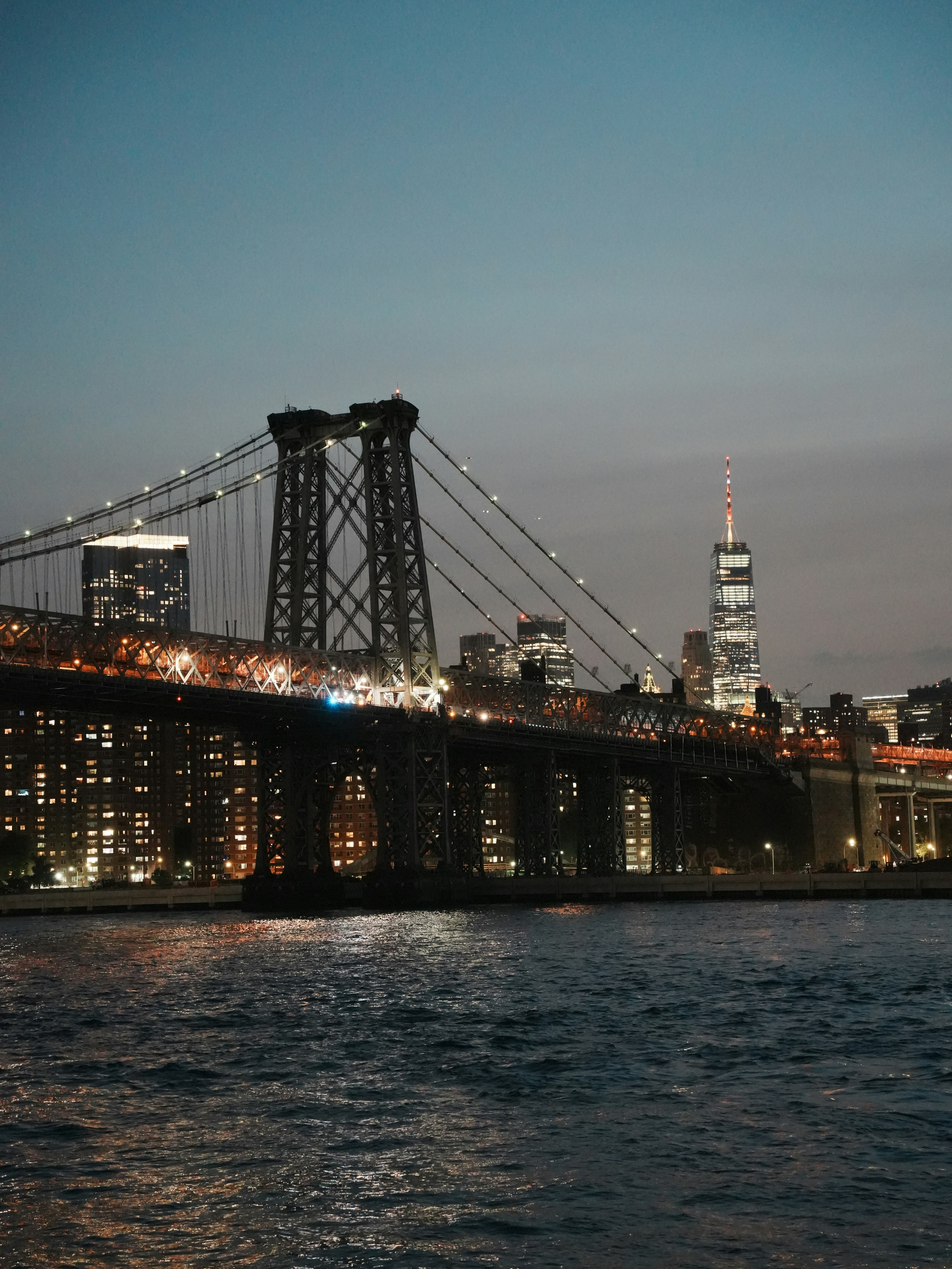 Williamsburg Bridge illuminated at dusk, with the skyline of New York City in the background featuring the Empire State Building.