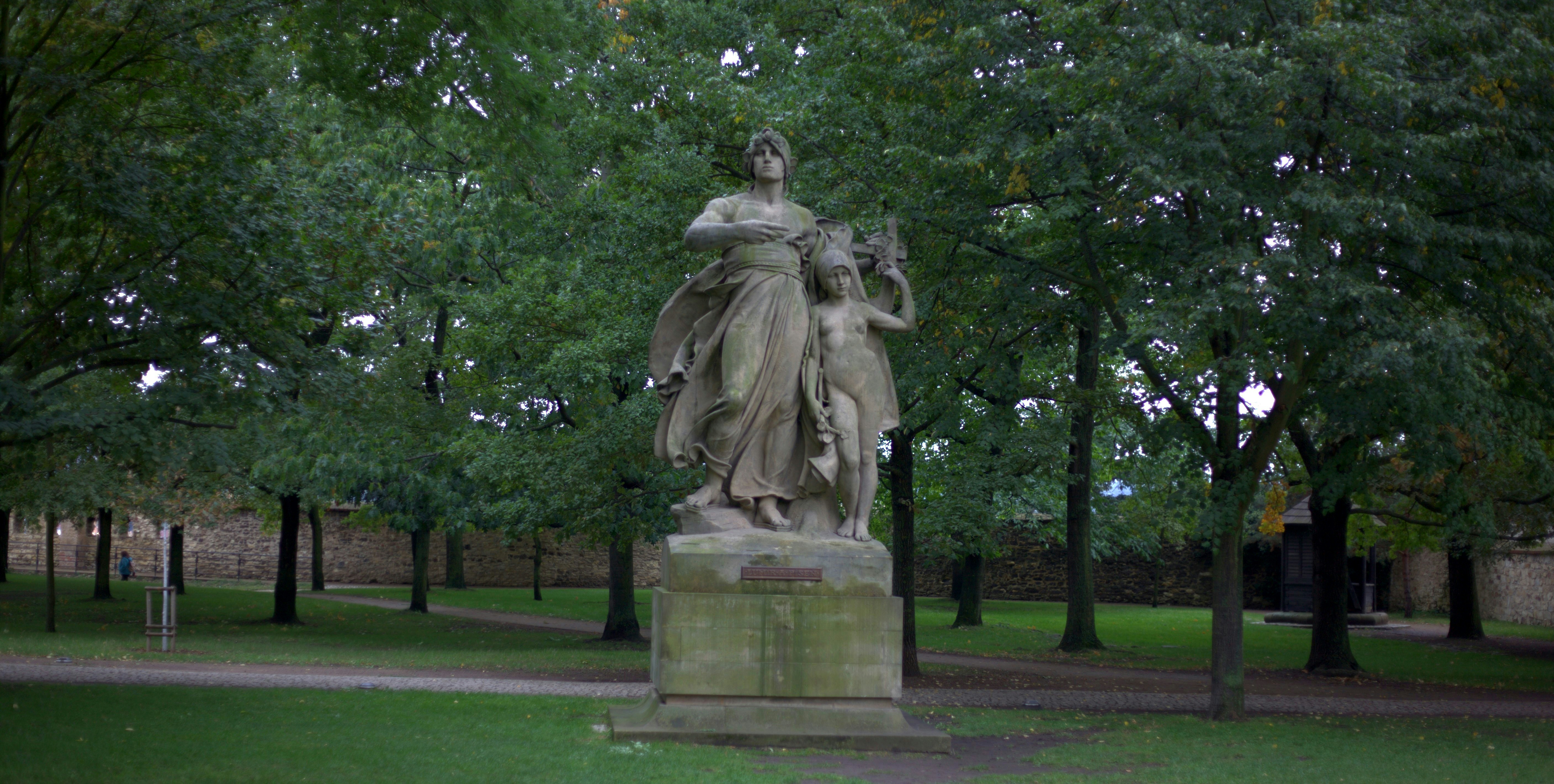 A majestic stone statue depicting a woman and child surrounded by lush greenery in a tranquil park setting.