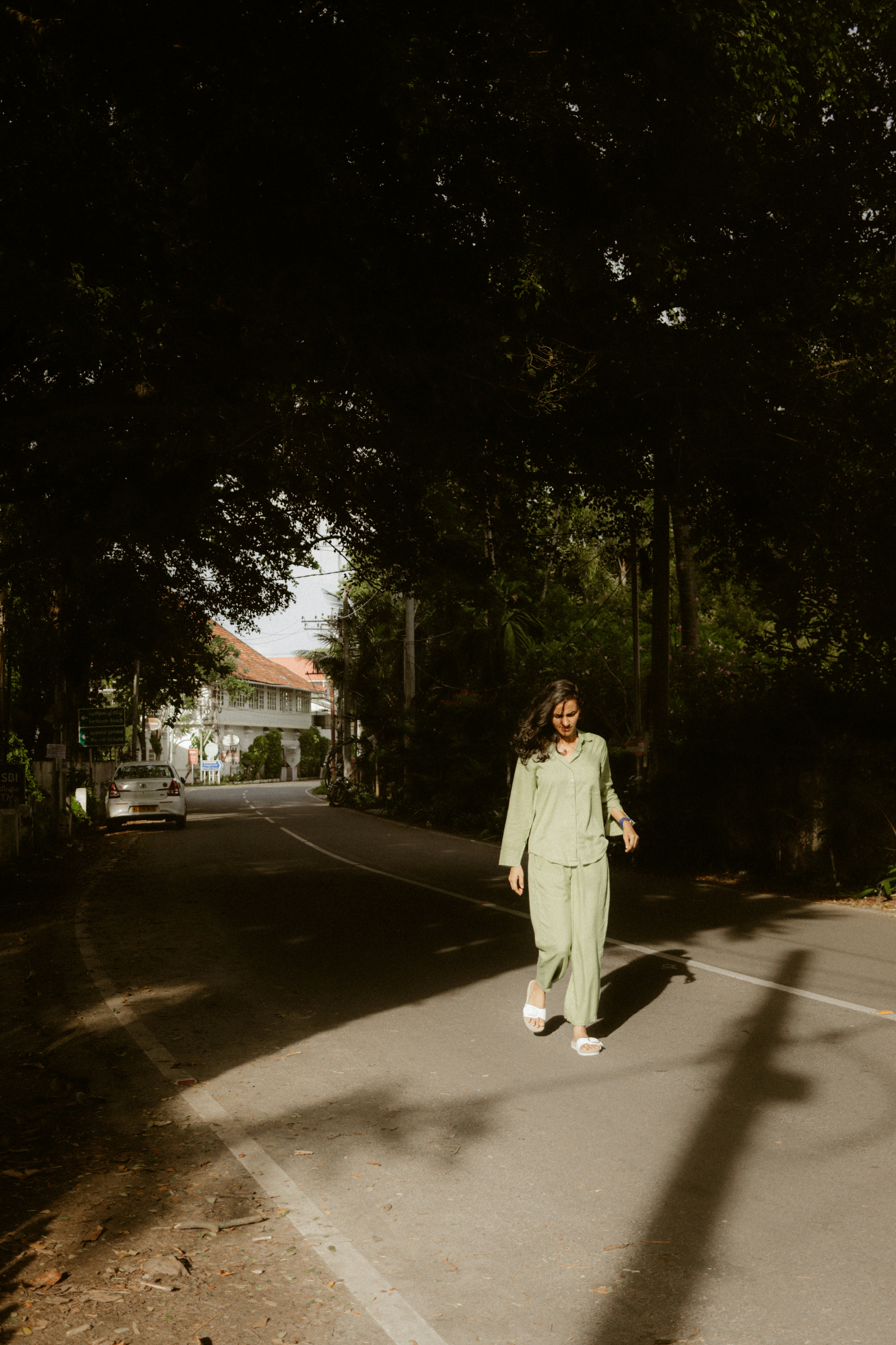 Woman in a light green outfit strolling along a sunlit road framed by lush trees, casting soft shadows on the pavement.
