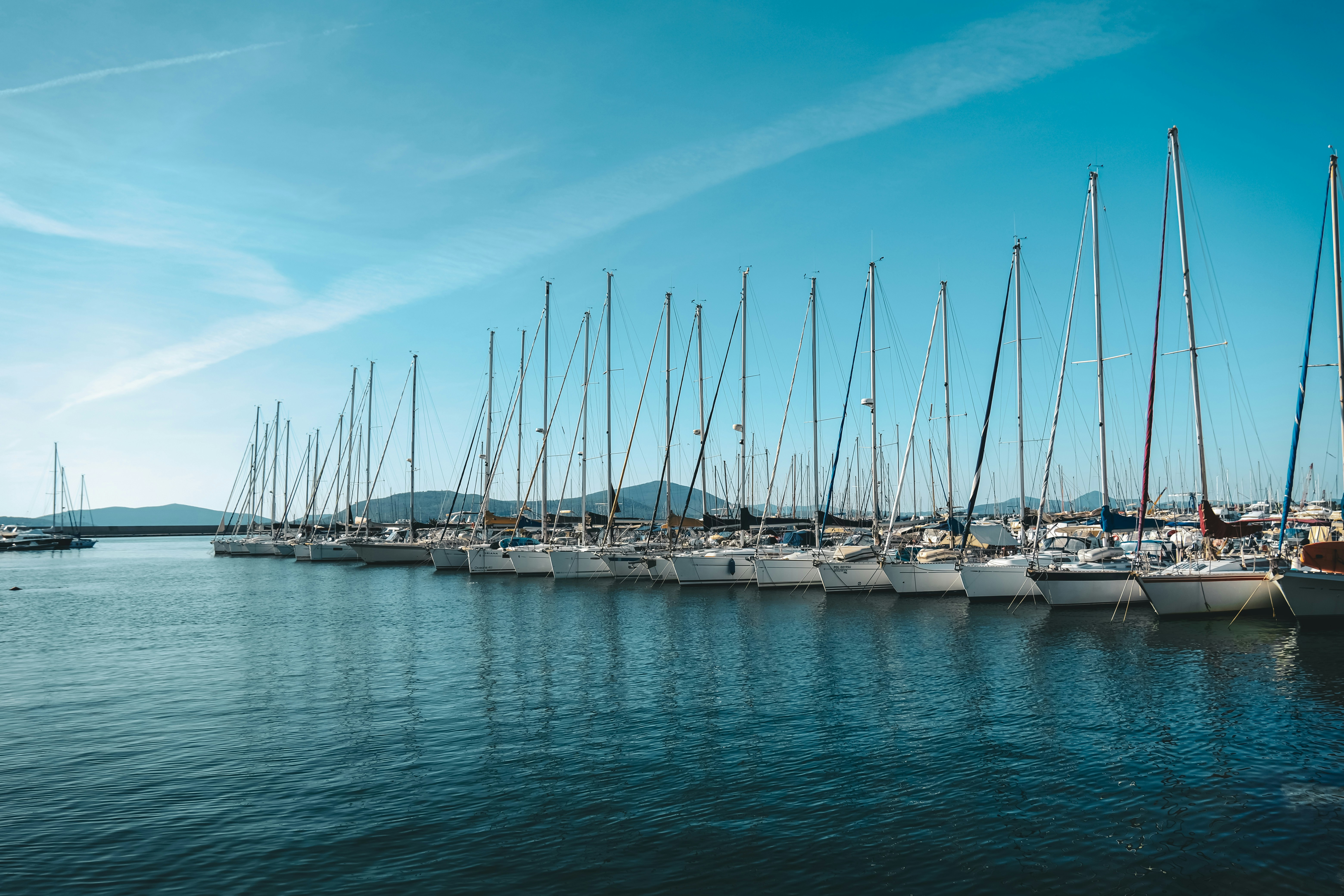 A row of sailboats anchored in a tranquil marina, reflecting in the calm waters under a clear blue sky.