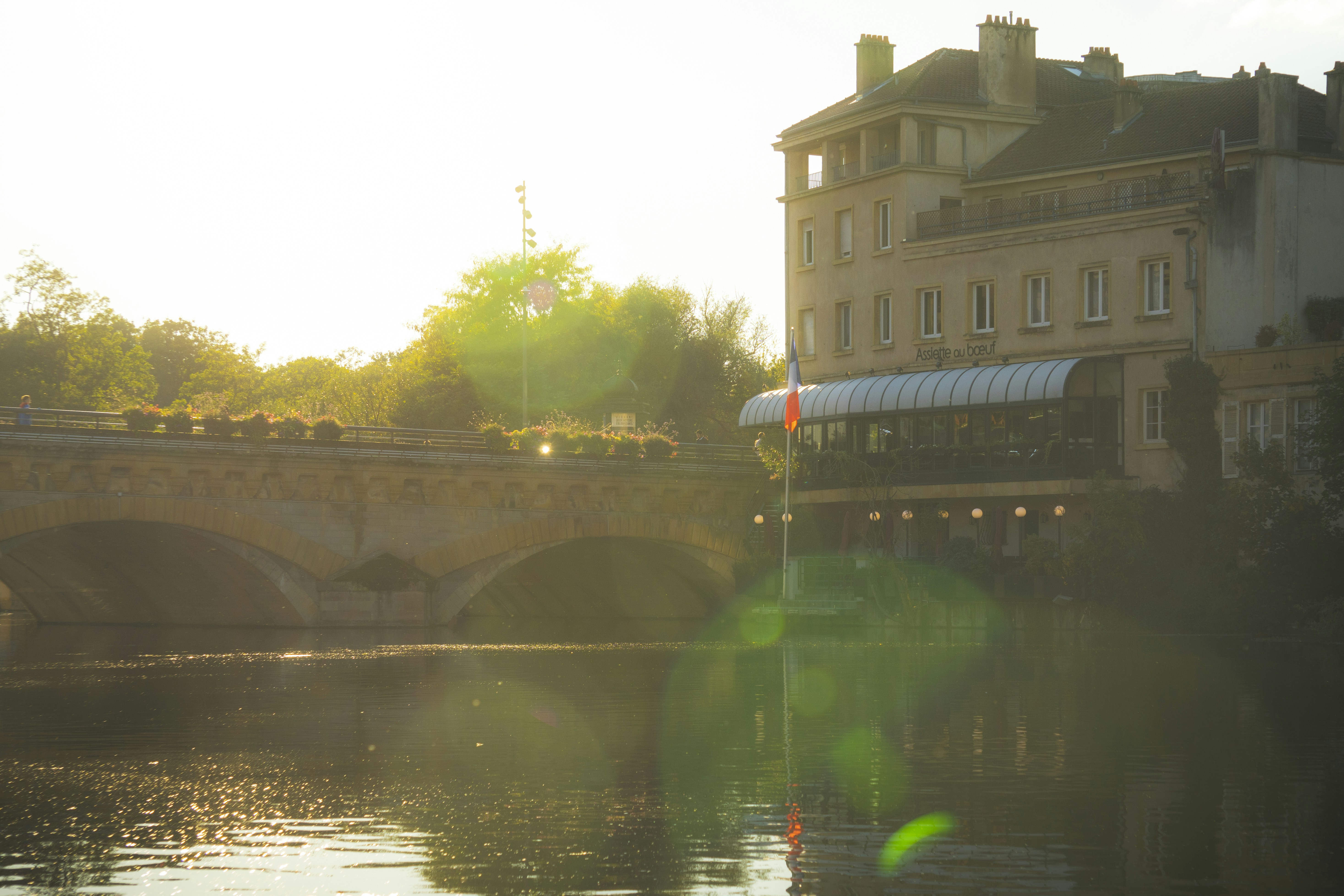 Historic stone bridge arching over a calm river, with a charming building adorned with flowers and a flag visible in the background. Sunlight casts a warm glow over the scene.