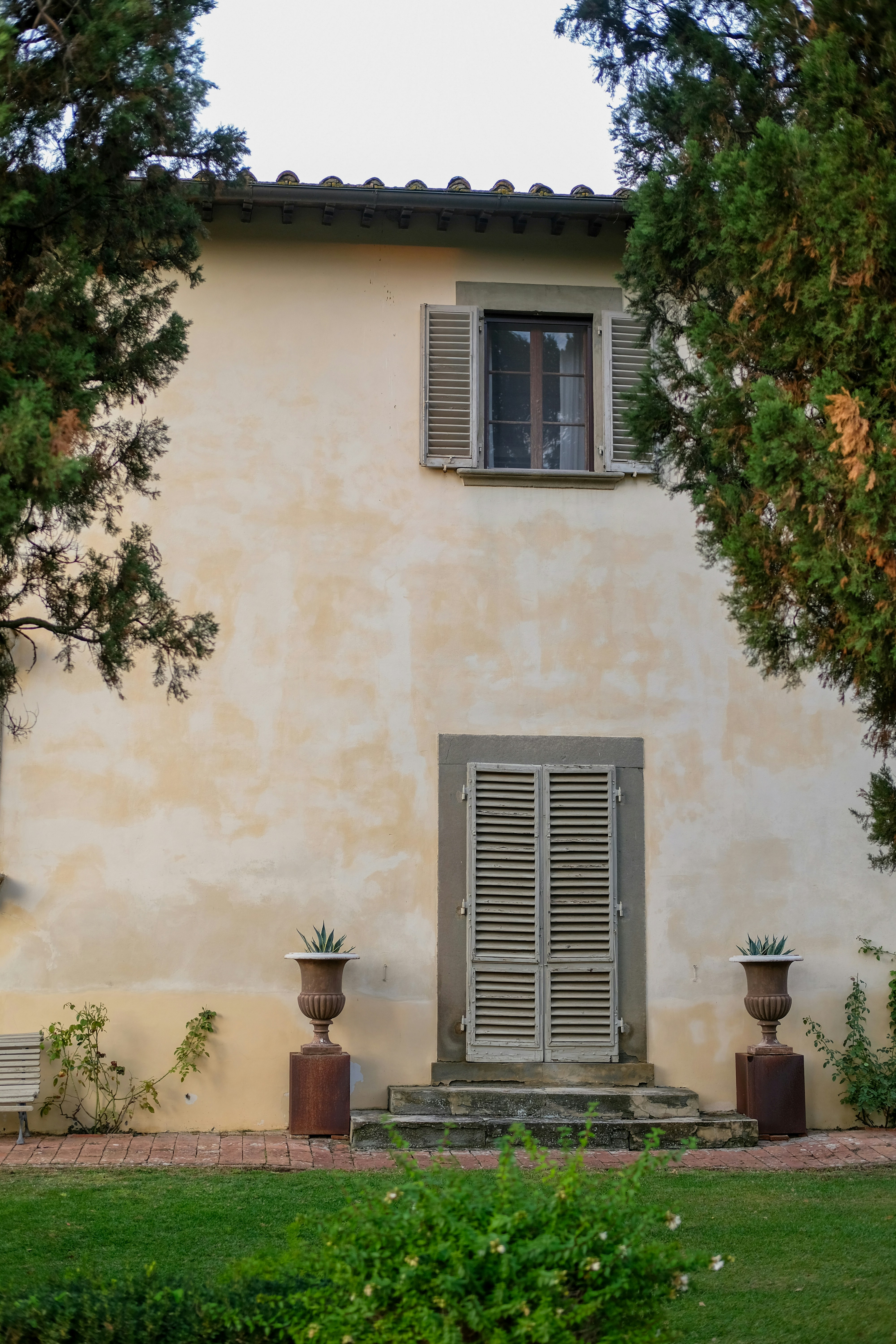 Charming facade of a rustic building framed by greenery, featuring a shuttered door and decorative pots. The soft colors evoke a serene atmosphere.