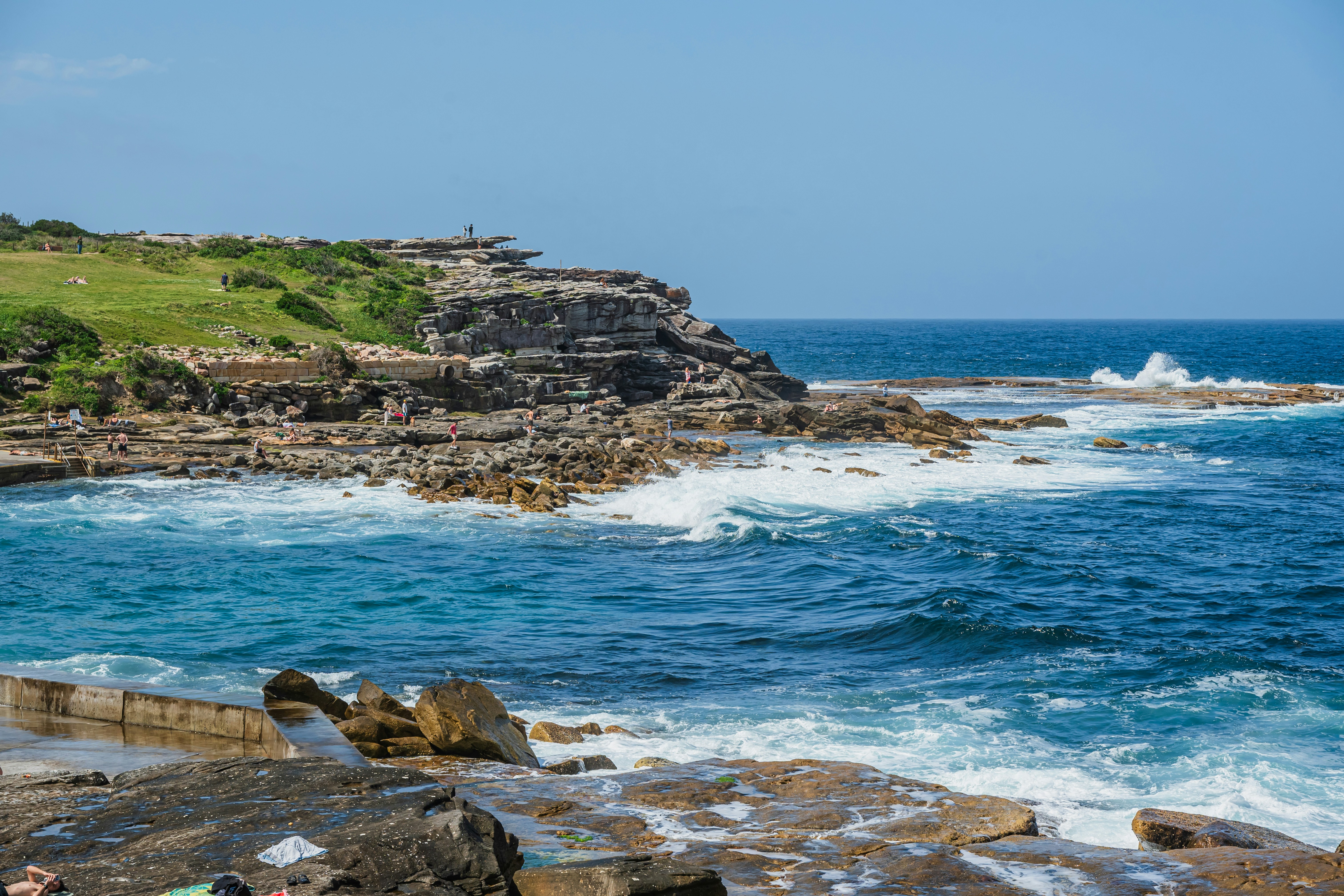 Rugged coastline meets vibrant blue waters, with gentle waves crashing against the rocks. Sunlit cliffs provide a backdrop to a serene seaside scene.