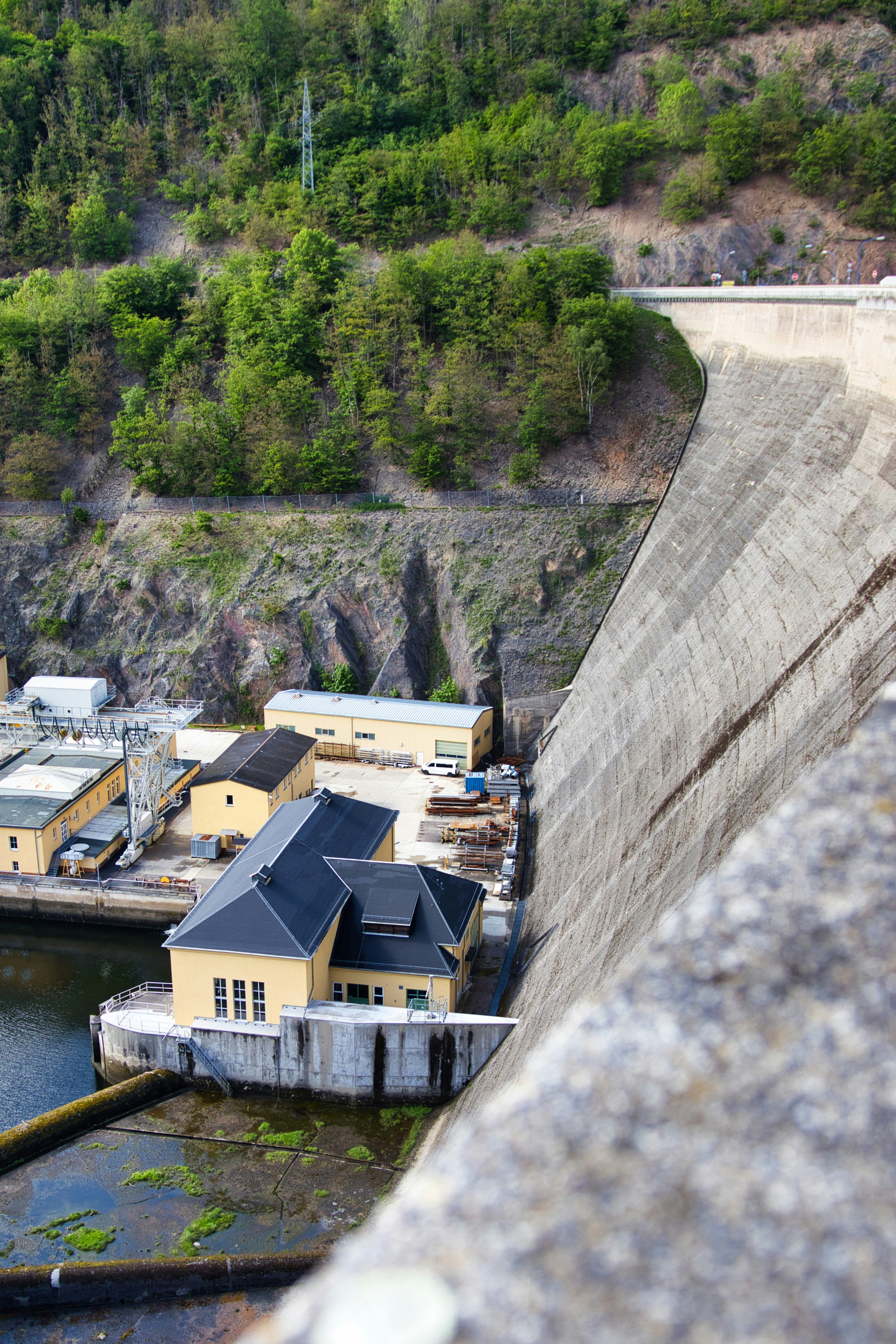 Aerial view of a dam with industrial buildings nestled at its base, surrounded by lush greenery and rocky terrain. The structure looms majestically over the water below.