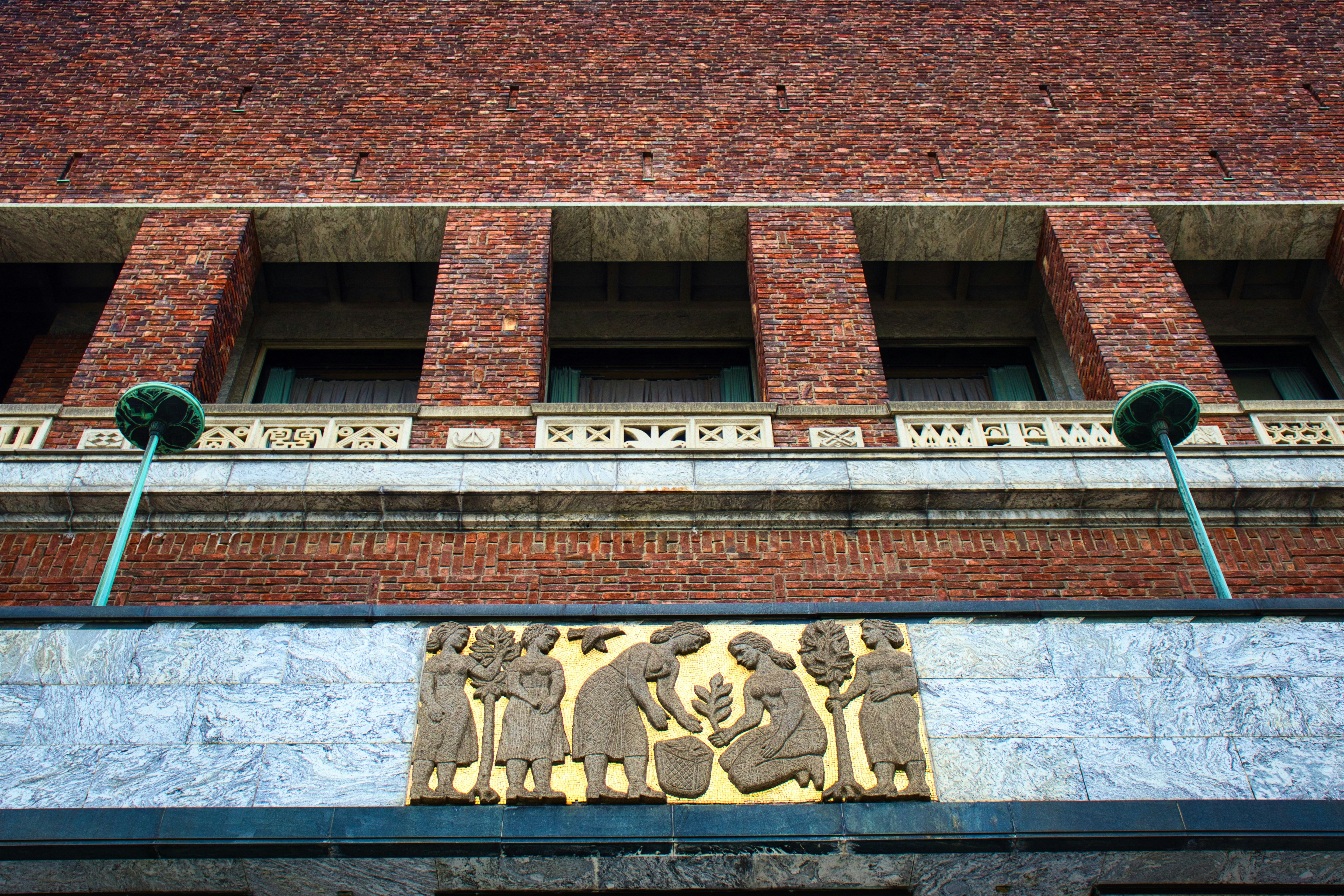 Architectural detail featuring a gold relief sculpture beneath a brick facade, showcasing human figures engaged in nature-themed activities.
