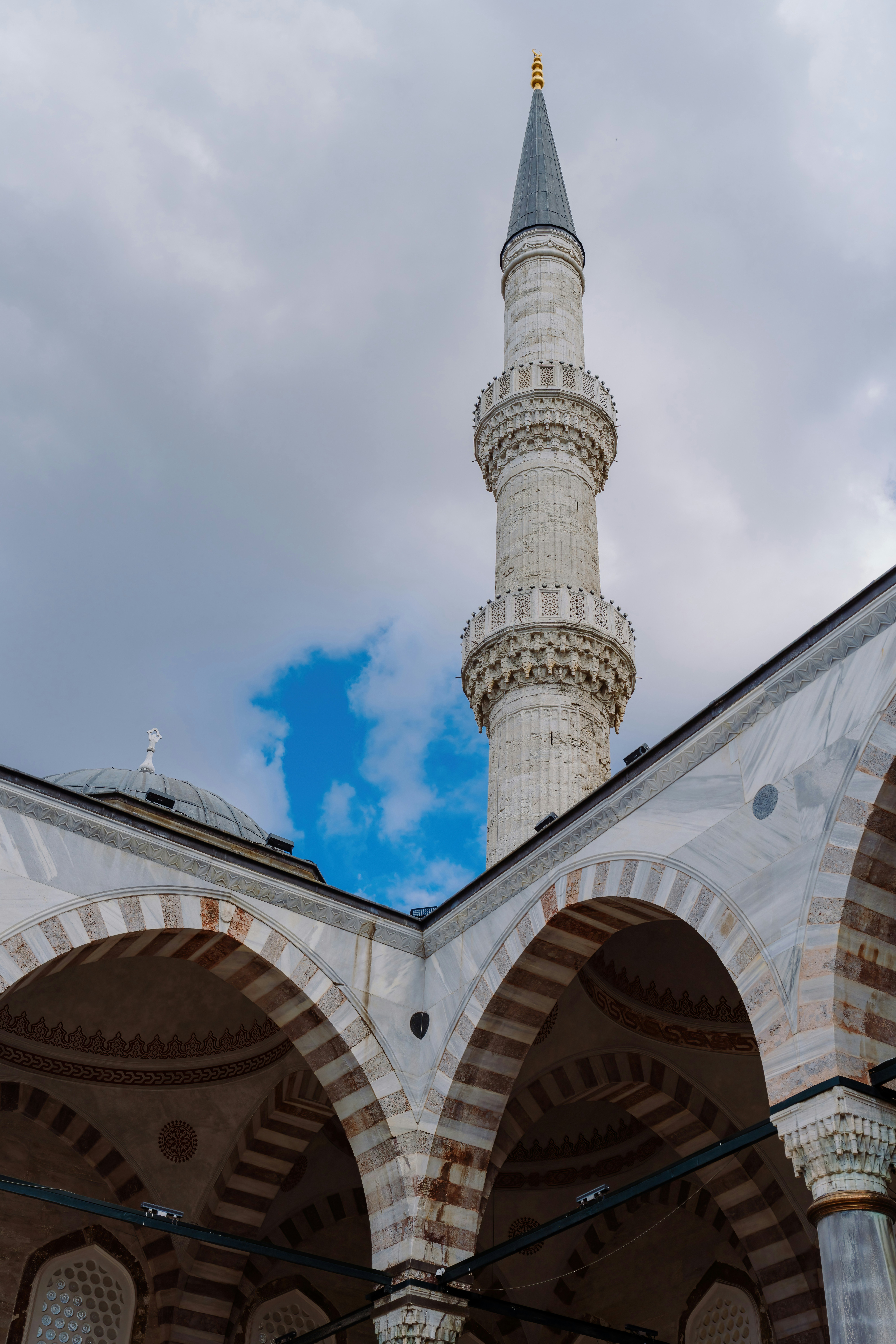 Elegant minaret rising above intricate arches of a mosque, framed by a dramatic sky. The architectural beauty reflects cultural heritage.