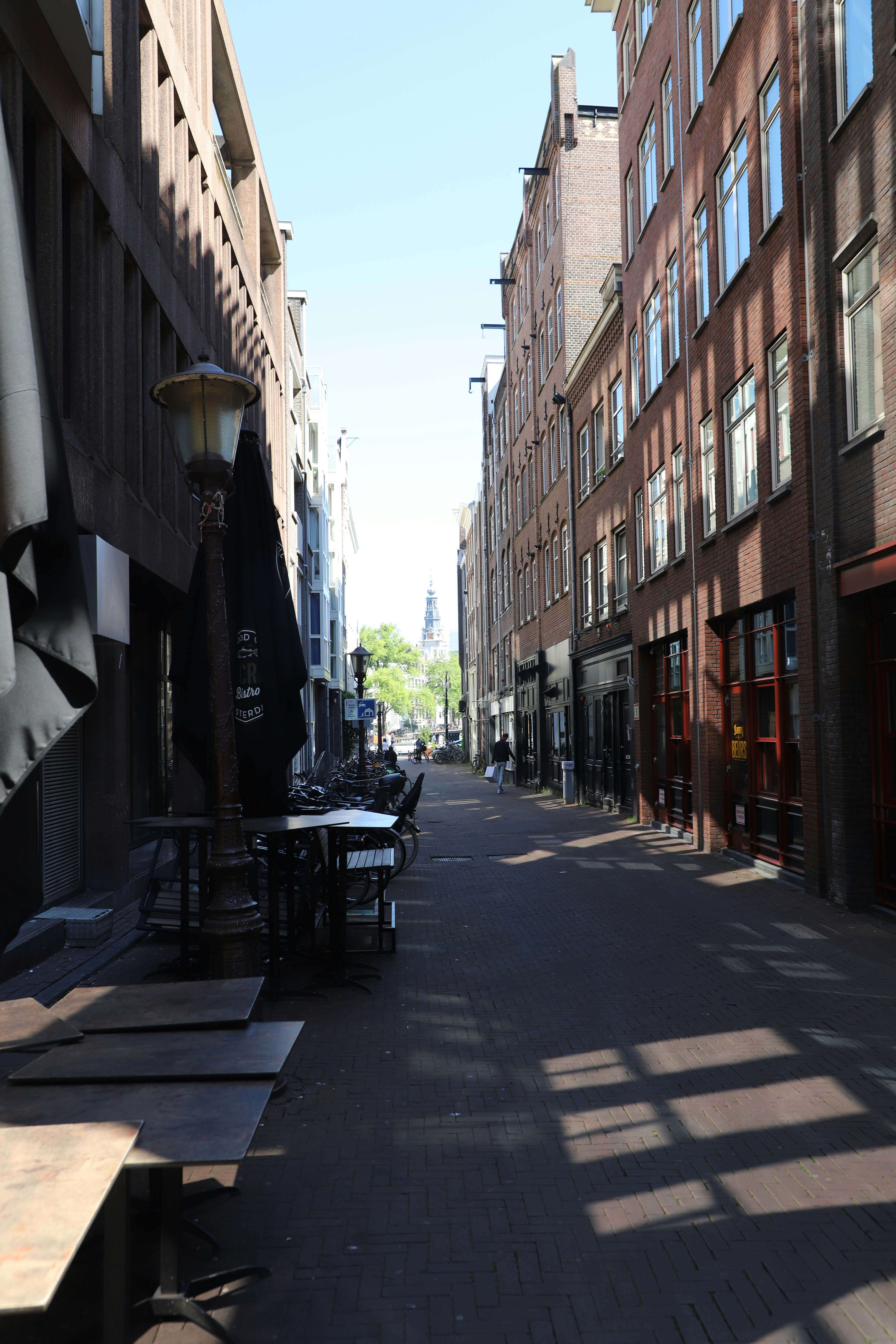 Sunlit alley lined with historic buildings, leading towards a distant tower. Tables and chairs hint at a lively café atmosphere.