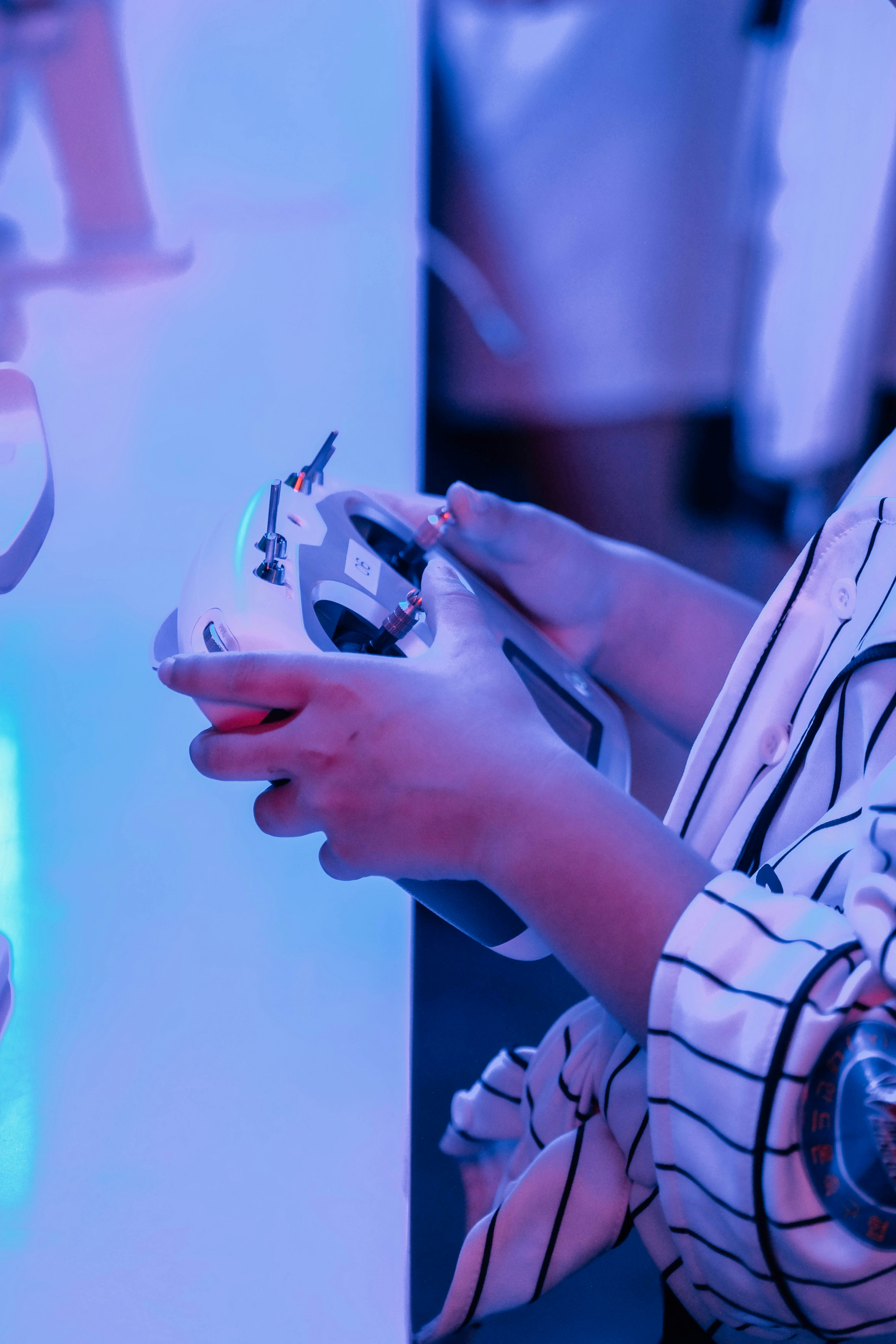 At the 2025 Drone World Cup in Jeonju, South Korea, the virtual reality drone soccer exhibition attracted enthusiastic participants eager to step into the future of the sport. Inside the specially designed booth, players donned VR headsets and gripped flight controllers, guiding their virtual drones across a digital arena that faithfully replicated the speed and intensity of real matches. Spectators gathered around the zone. | Hands holding a remote control with blue lighting