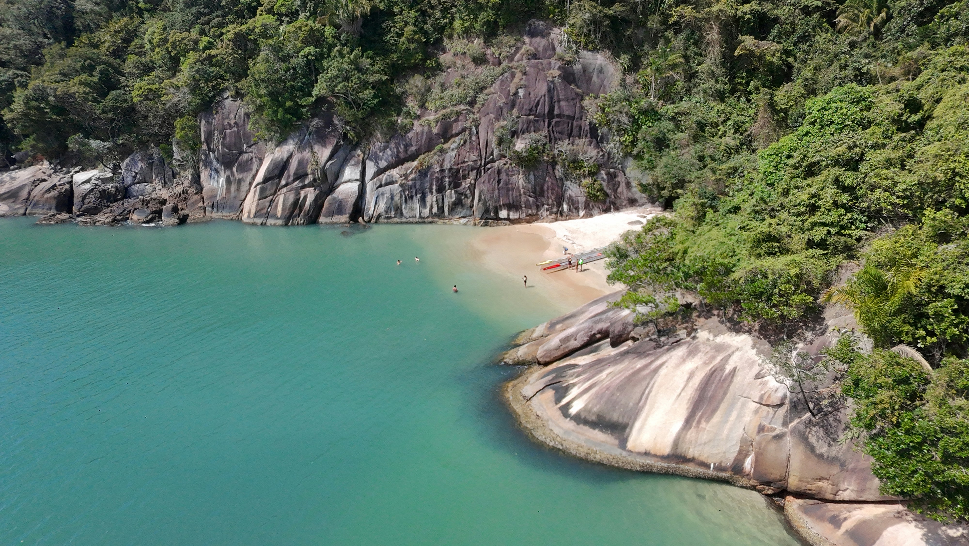 A tranquil beach nestled between rocky cliffs and lush greenery, with a few people enjoying the serene waters. The scene captures the essence of a secluded paradise.