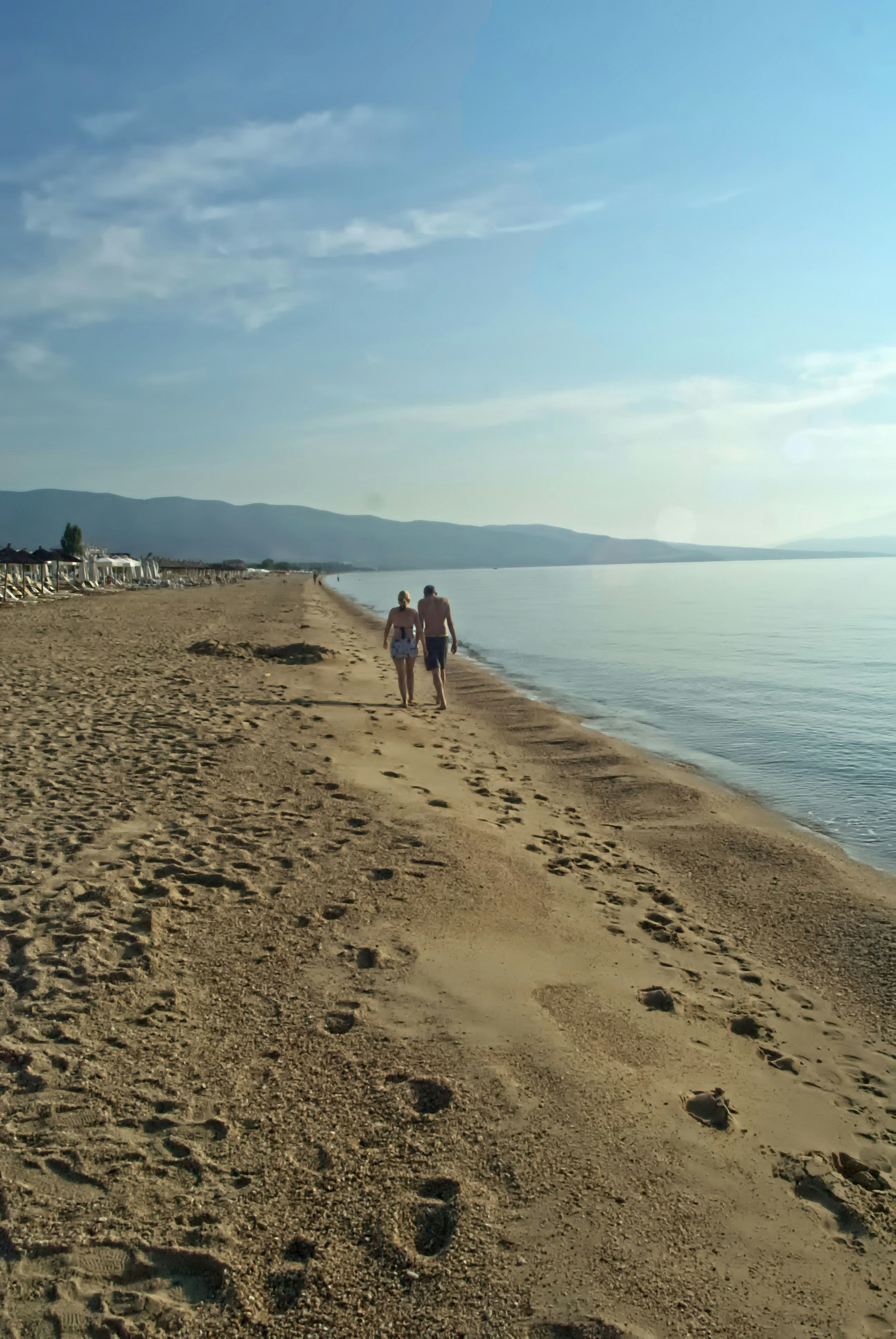 Two individuals walking along a sandy beach, with gentle waves lapping at the shore under a clear blue sky.