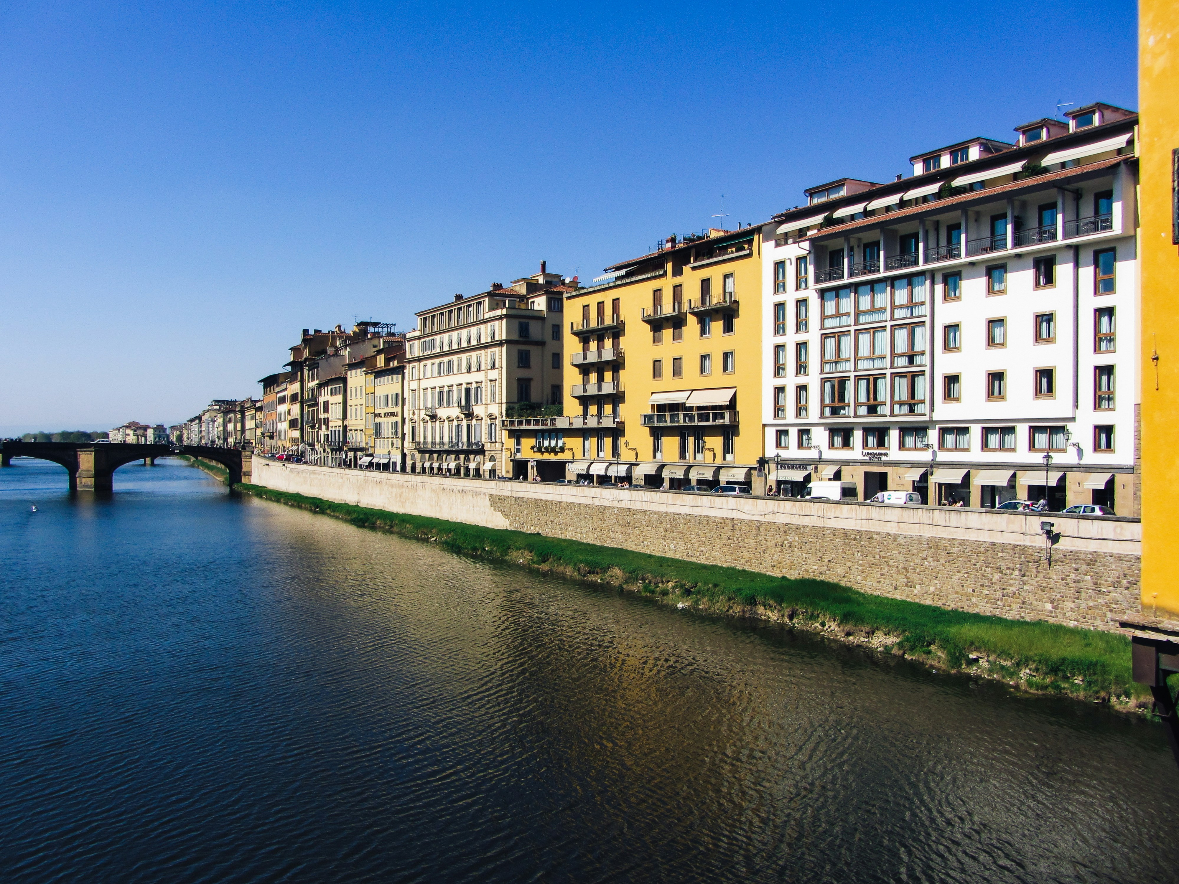 Along the Arno, view from Ponte alla Carraia | Buildings line a river with a bridge in the distance.