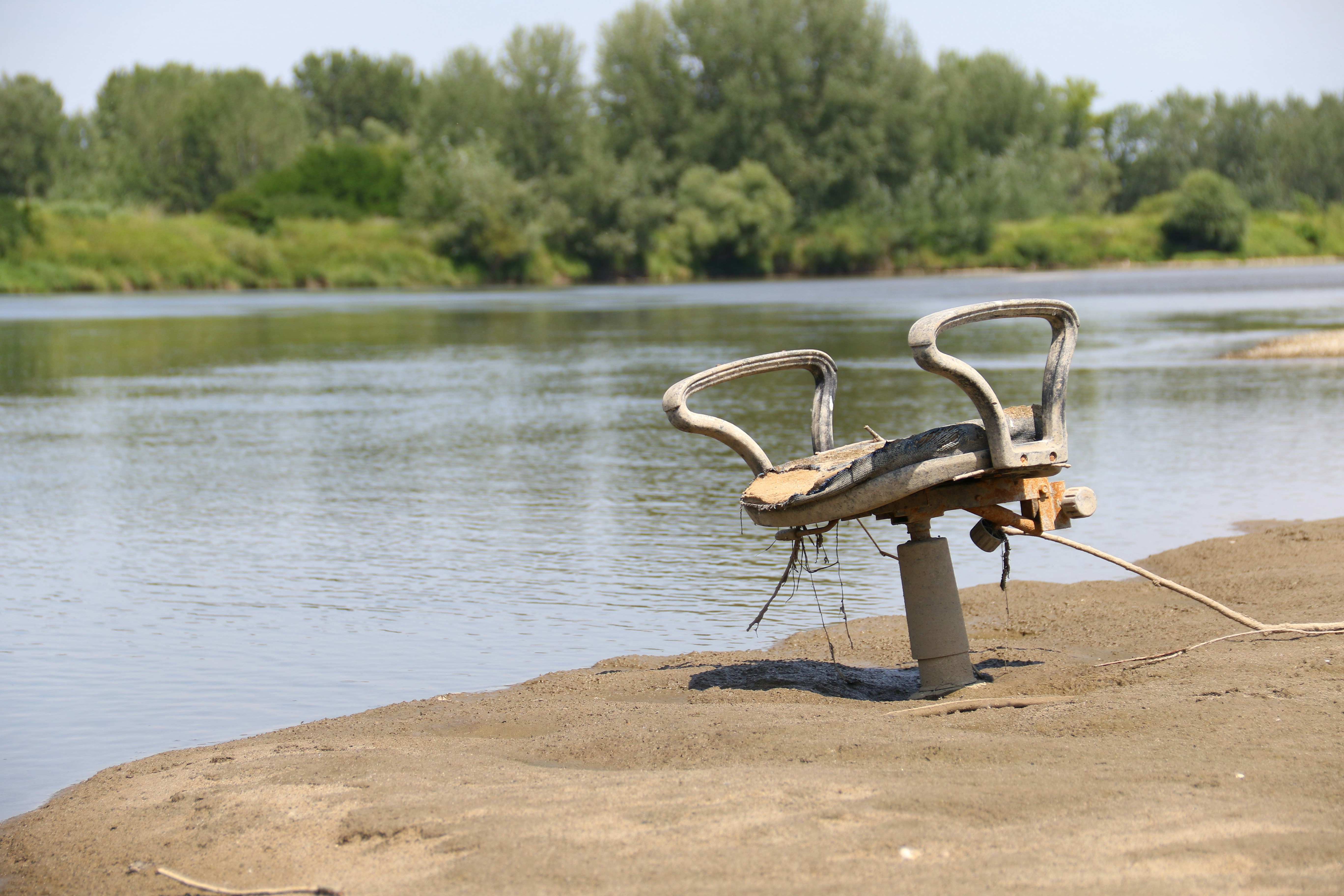 An abandoned office chair partially buried in sand near a tranquil river, surrounded by lush greenery.