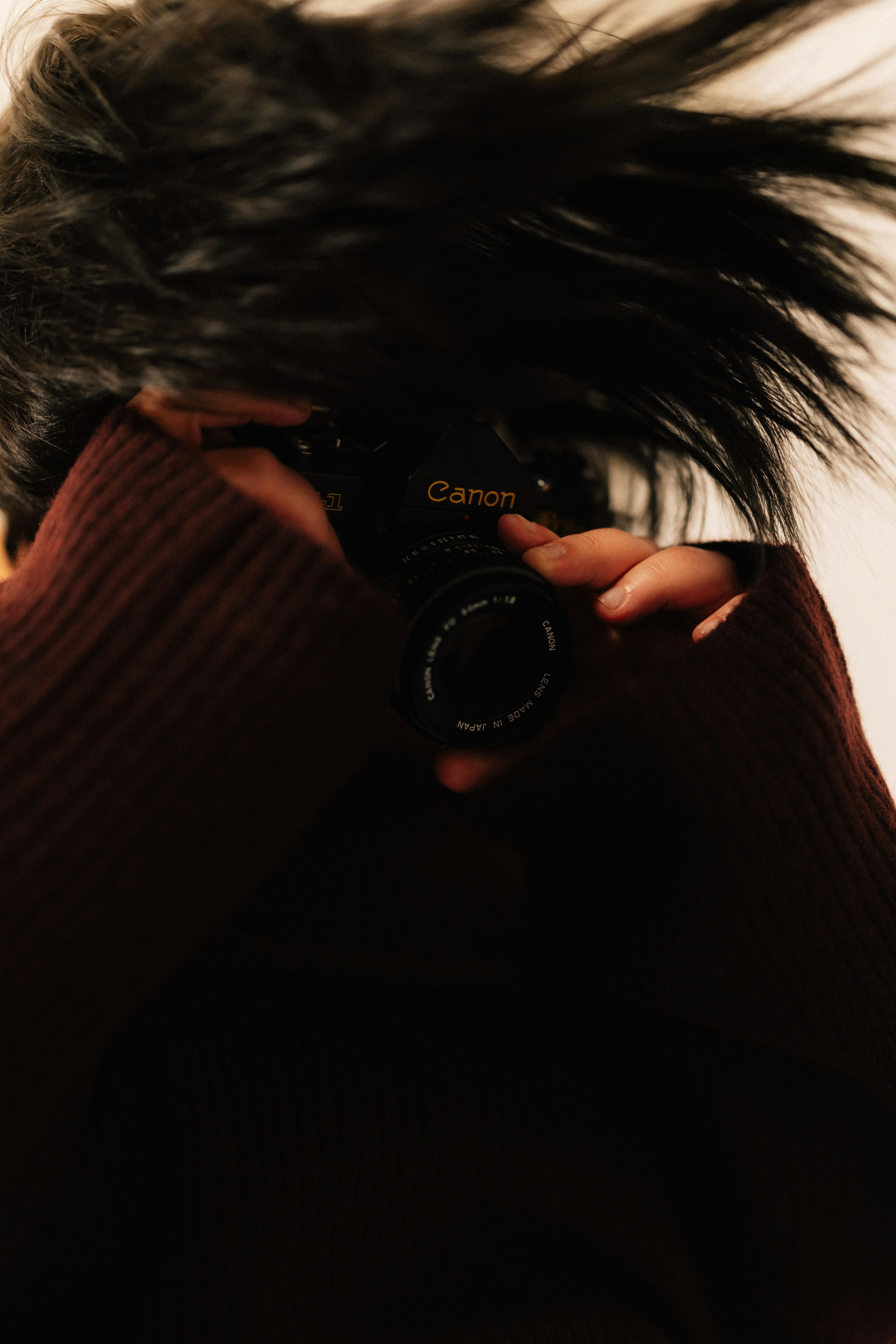 Person holding a Canon camera, partially obscured by their long hair, dressed in a cozy sweater. The scene evokes a sense of intimacy and creativity.