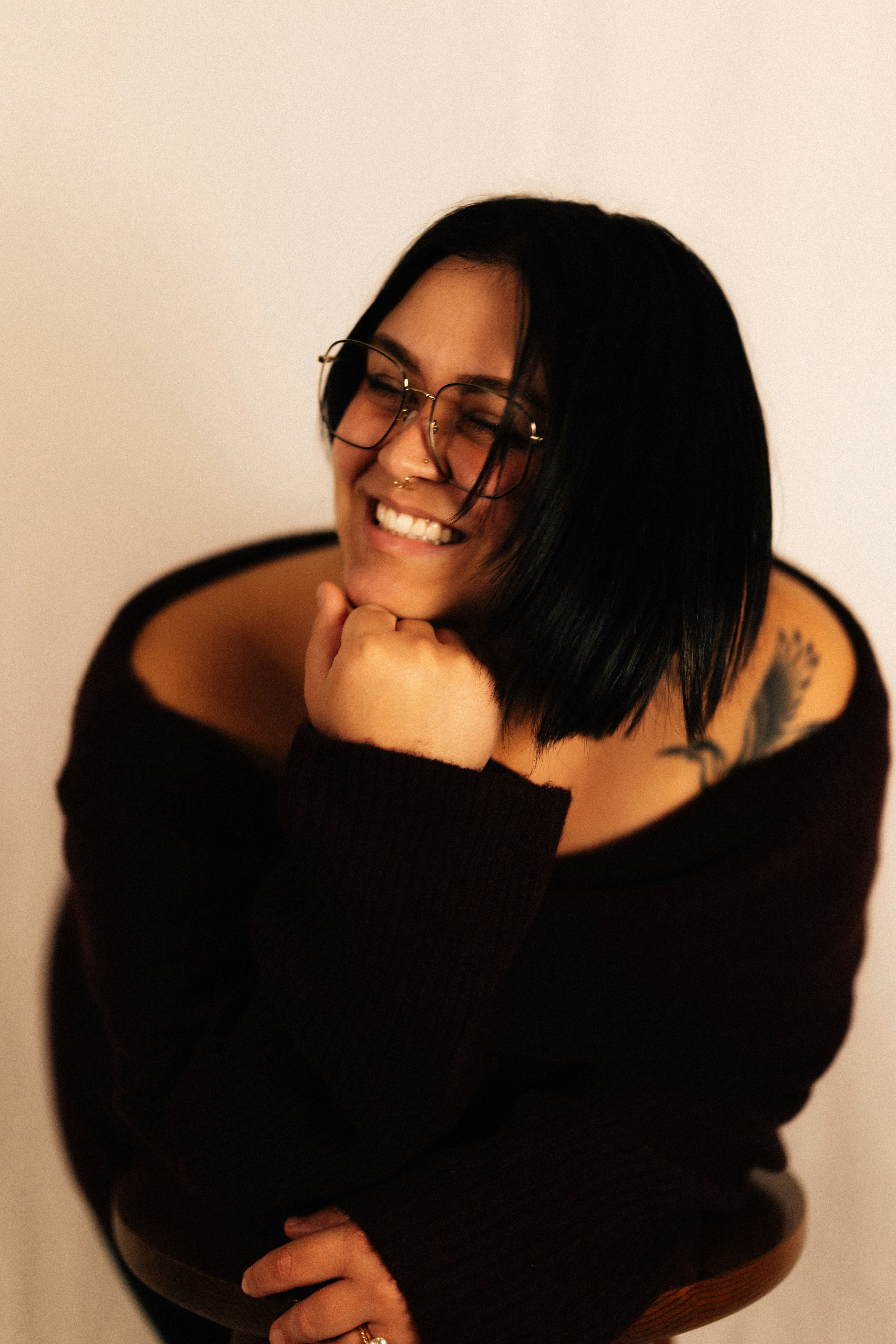 A smiling individual with short black hair and glasses poses comfortably in a dark oversized sweater, showcasing a tattoo on their shoulder against a soft, neutral backdrop.