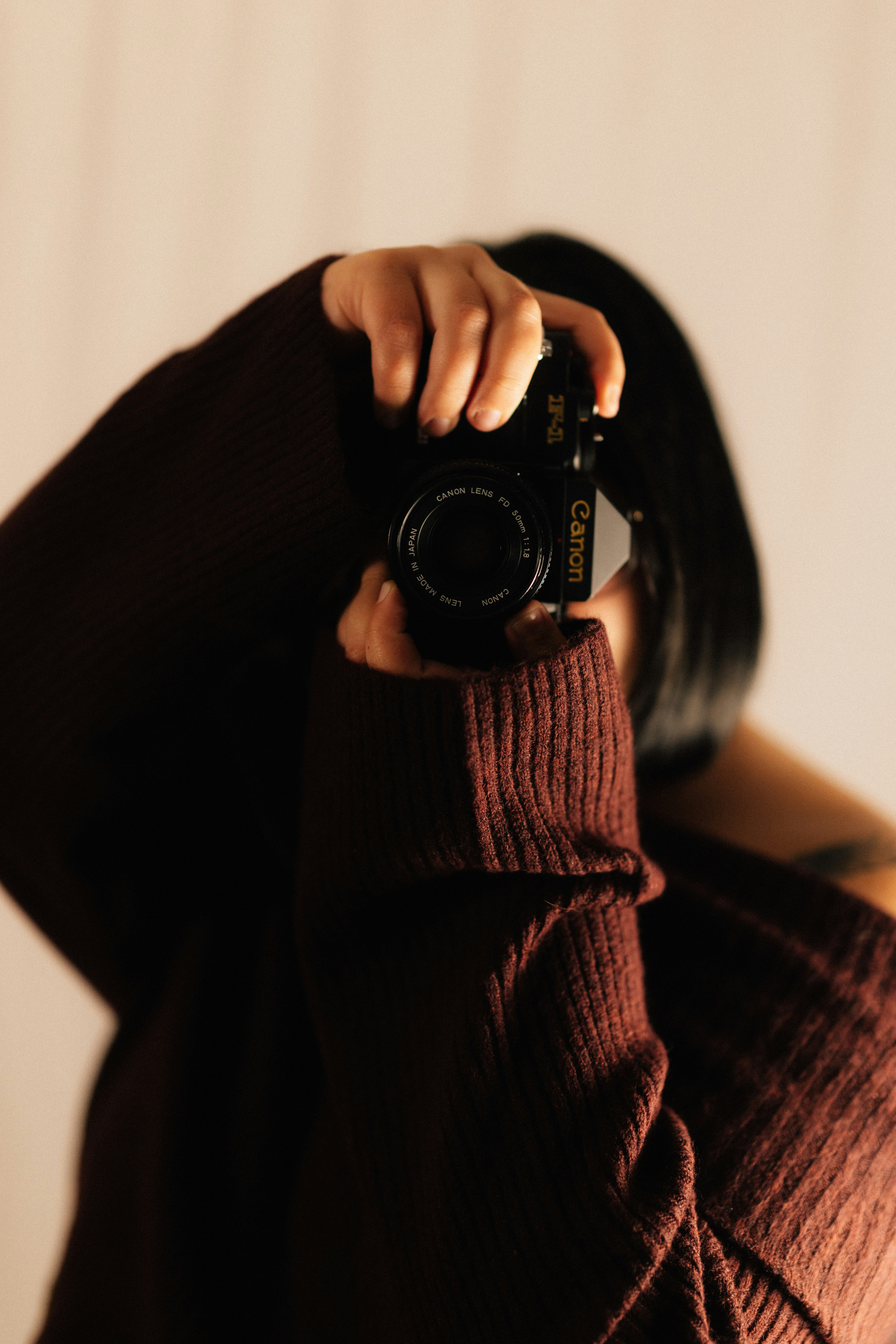 A person in a cozy maroon sweater holds a Canon camera, capturing a moment in a softly lit environment.