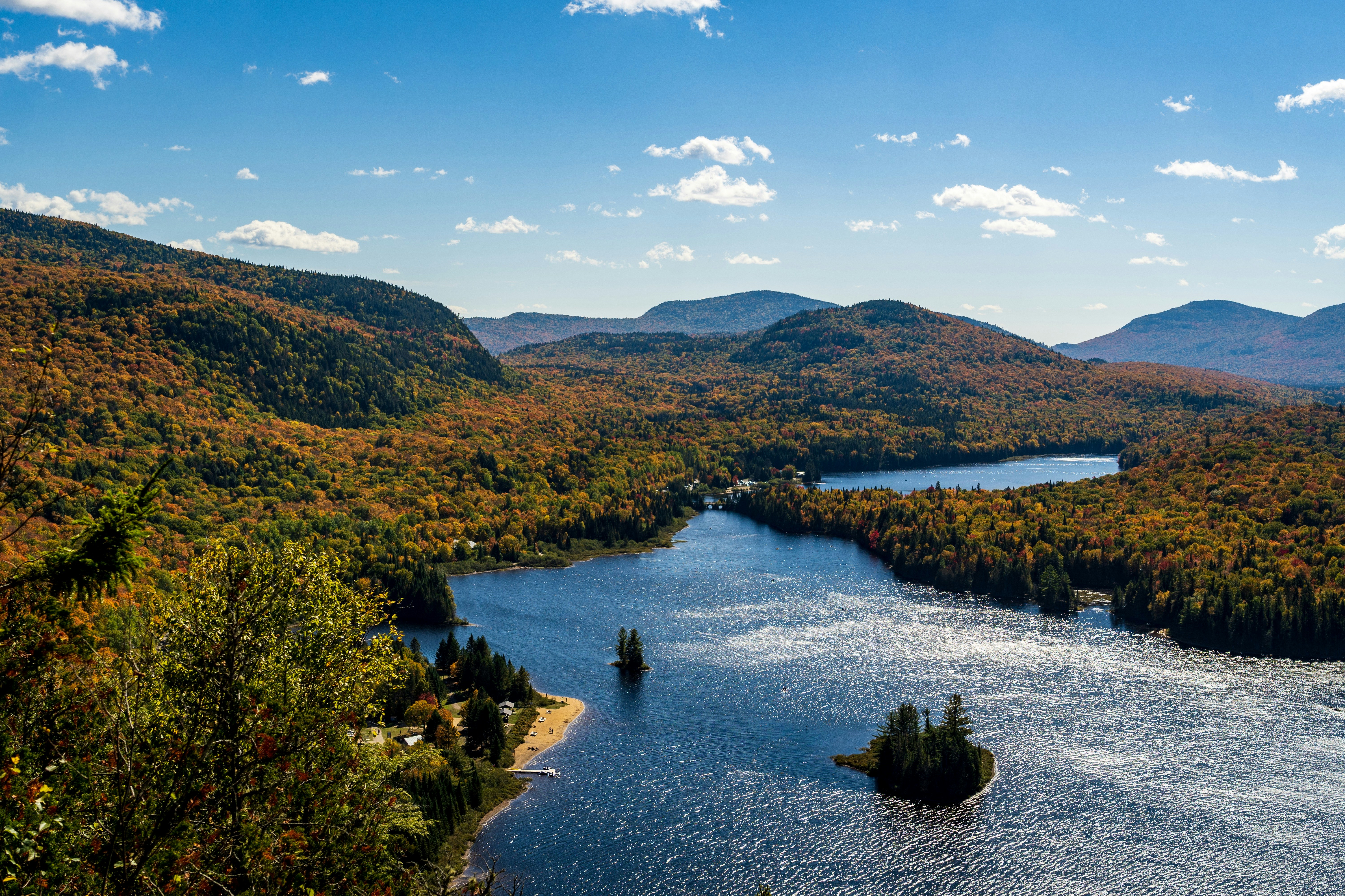 Vibrant autumn foliage blankets the hills surrounding a serene lake, reflecting the clear blue sky and scattered clouds.