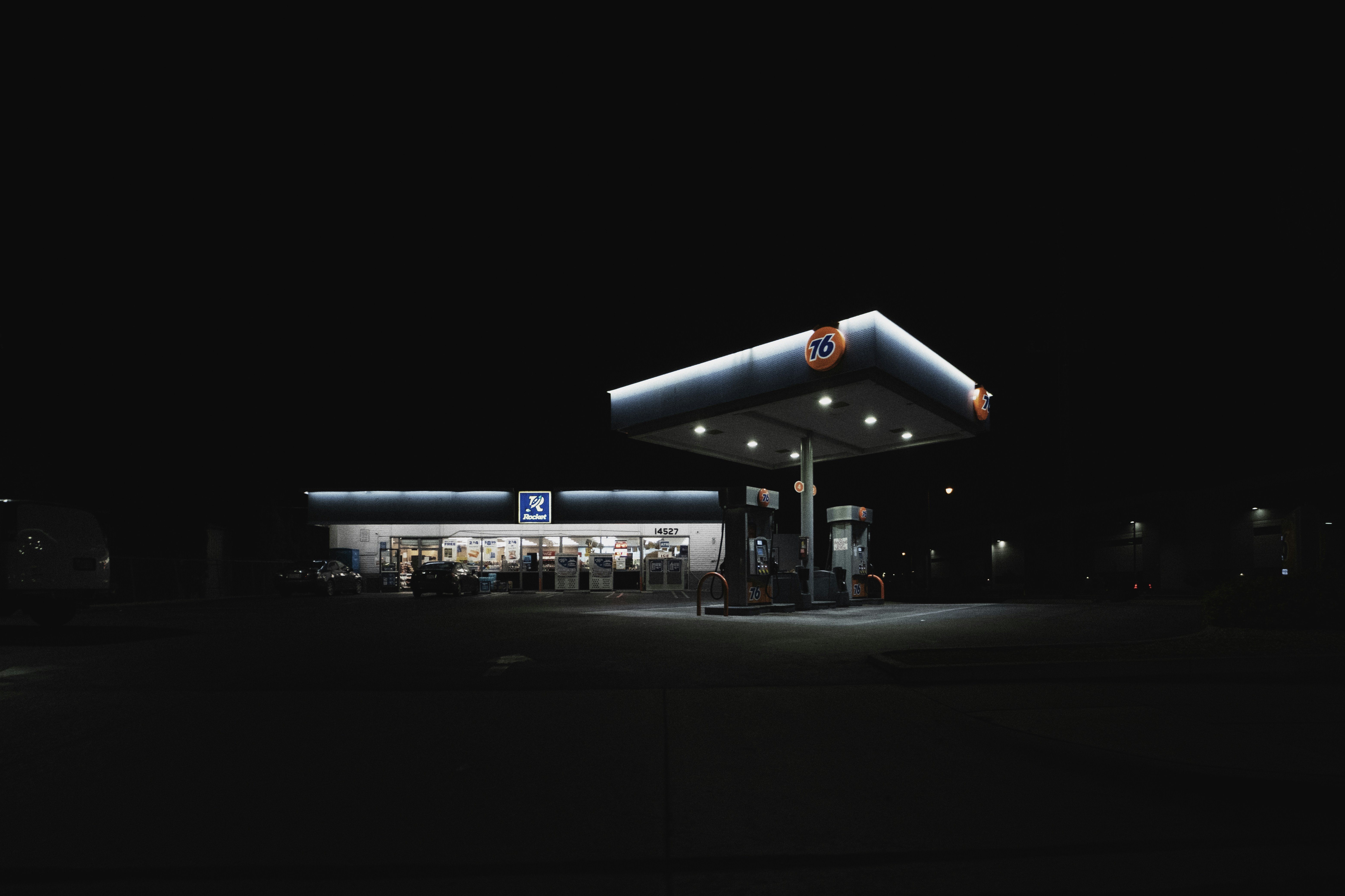 A gas station illuminated against the night sky, showcasing its bright signage and pumps. The scene captures the quiet stillness of late-night refueling.