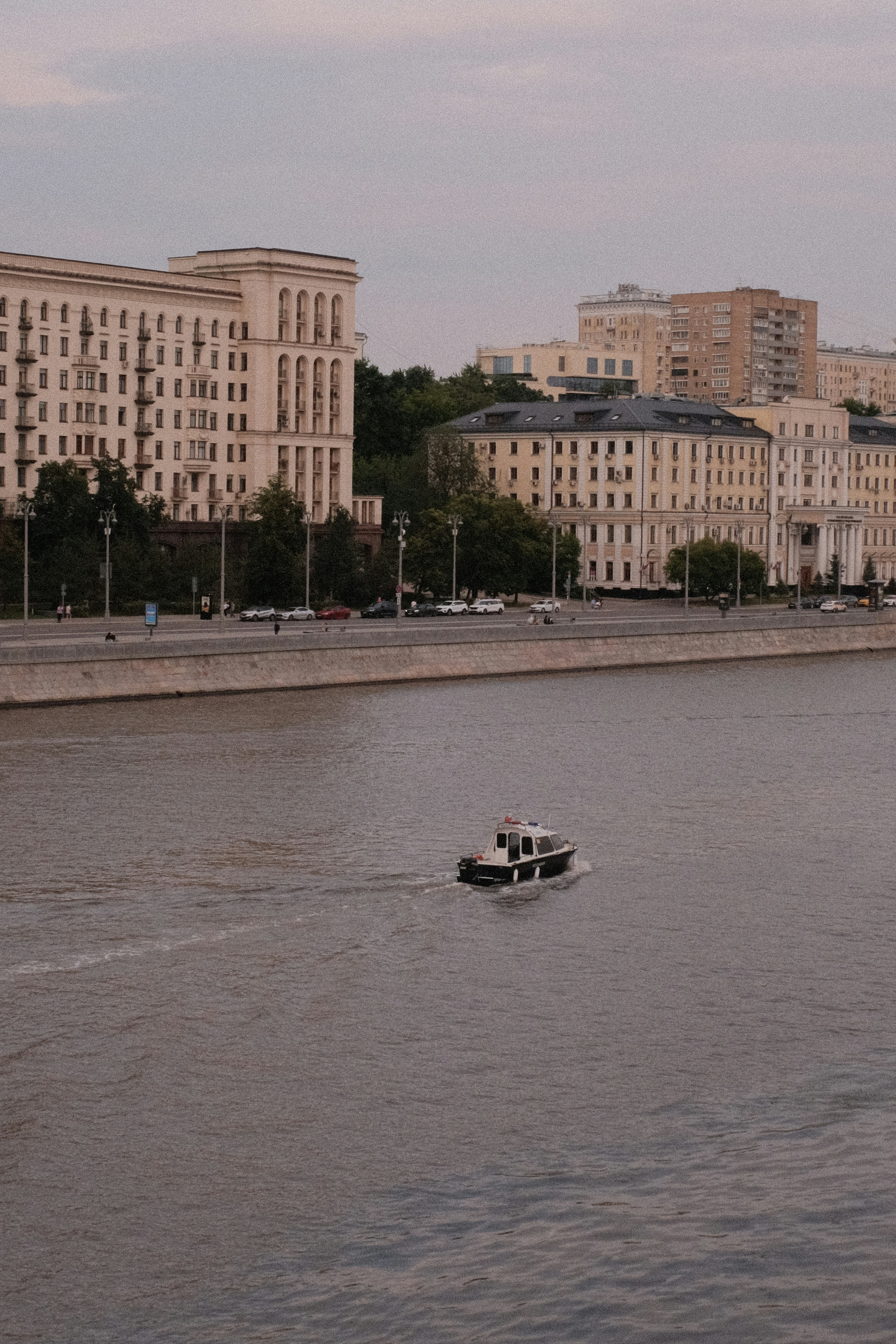 A boat glides through the calm waters of a river, framed by historic buildings along the bank. The tranquil scene captures the essence of urban life by the water.