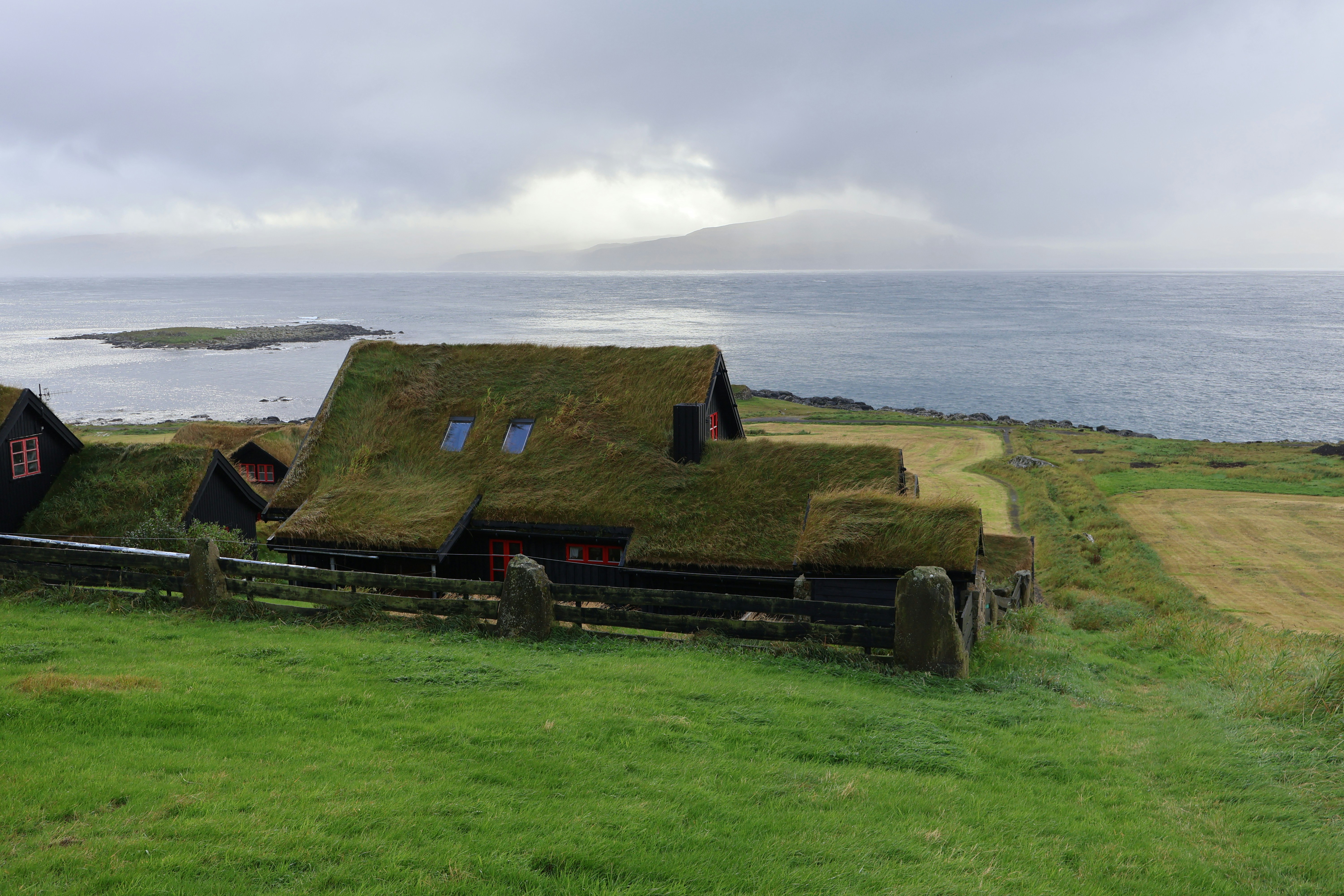 Traditional grass-roofed houses nestled on a lush hillside overlooking a tranquil sea, with distant islands shrouded in mist.