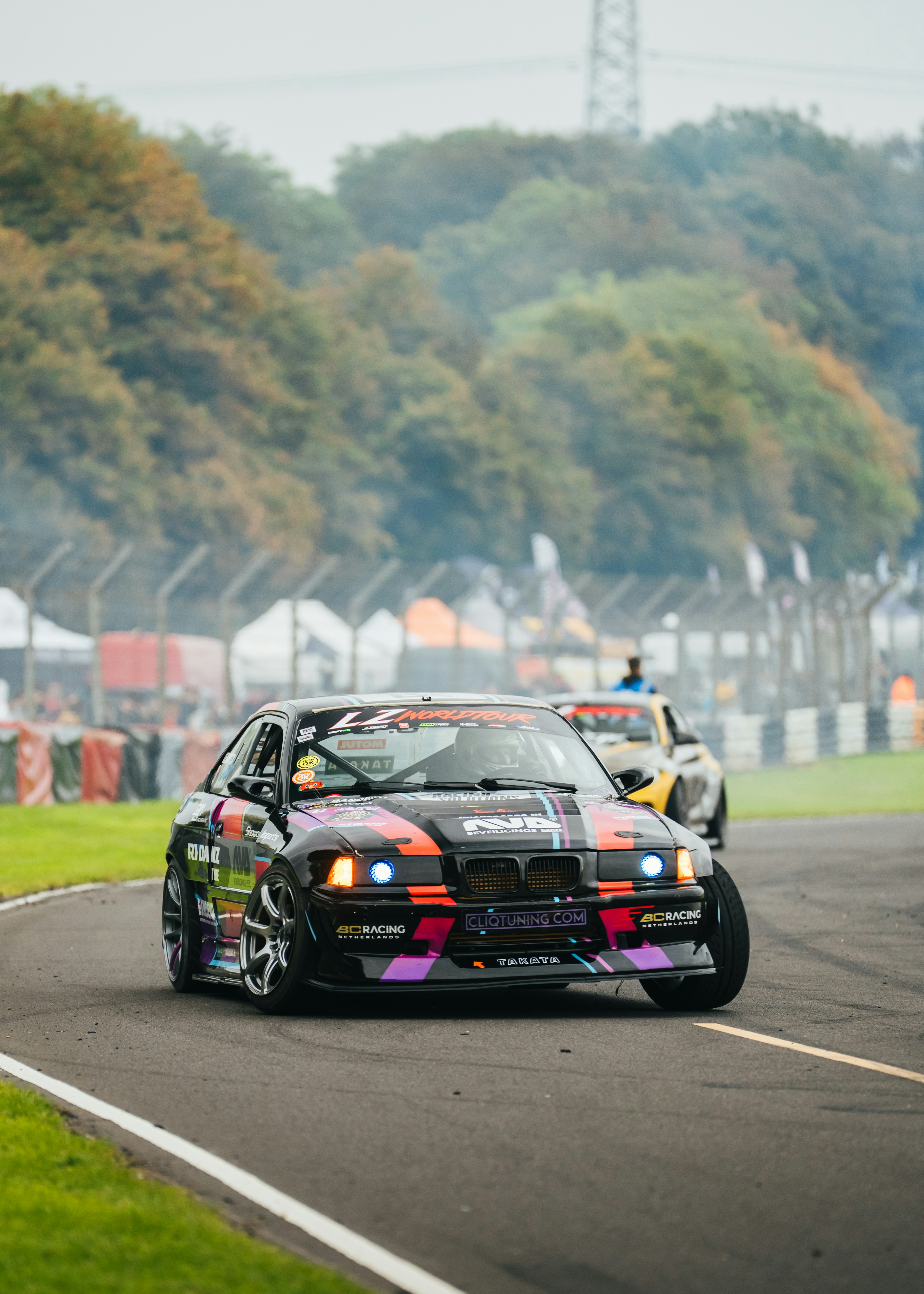 A vibrant drift car navigating a racetrack, surrounded by a backdrop of trees and spectators. The tire smoke adds to the thrilling atmosphere.