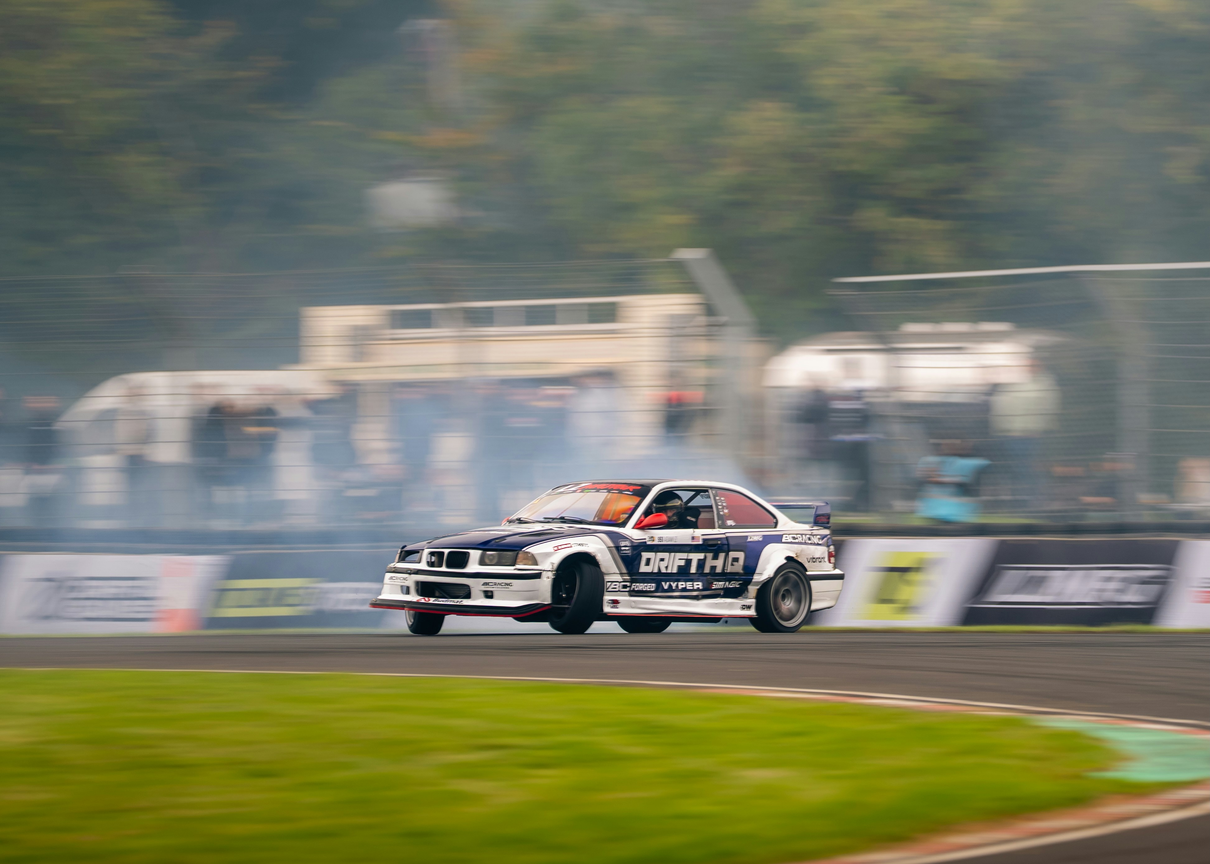 A drifting car expertly maneuvers around a track, surrounded by a haze of smoke and an engaged crowd. The scene captures the thrill of motorsport in action.