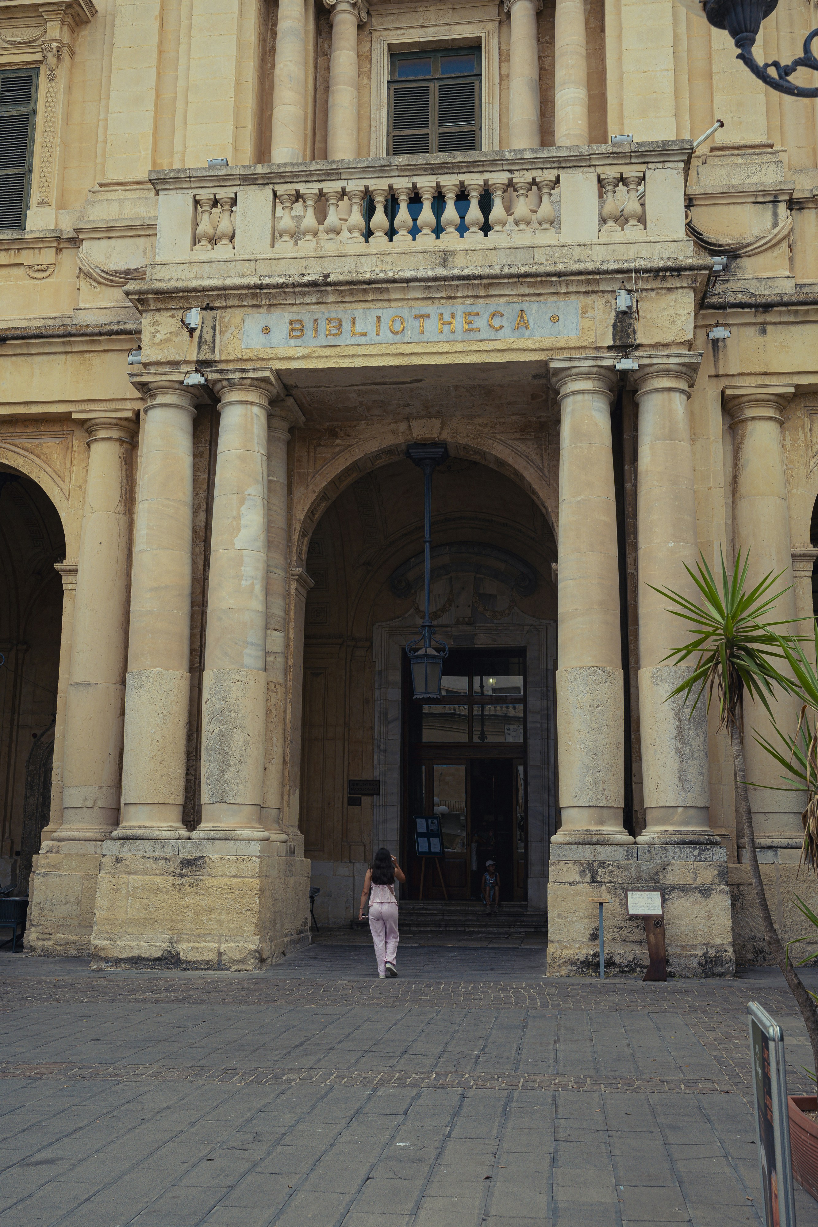 A woman approaches the grand entrance of a historic library adorned with classical columns and the inscription 'BIBLIOTHECA.' Lush greenery adds a touch of nature to the architectural scene.