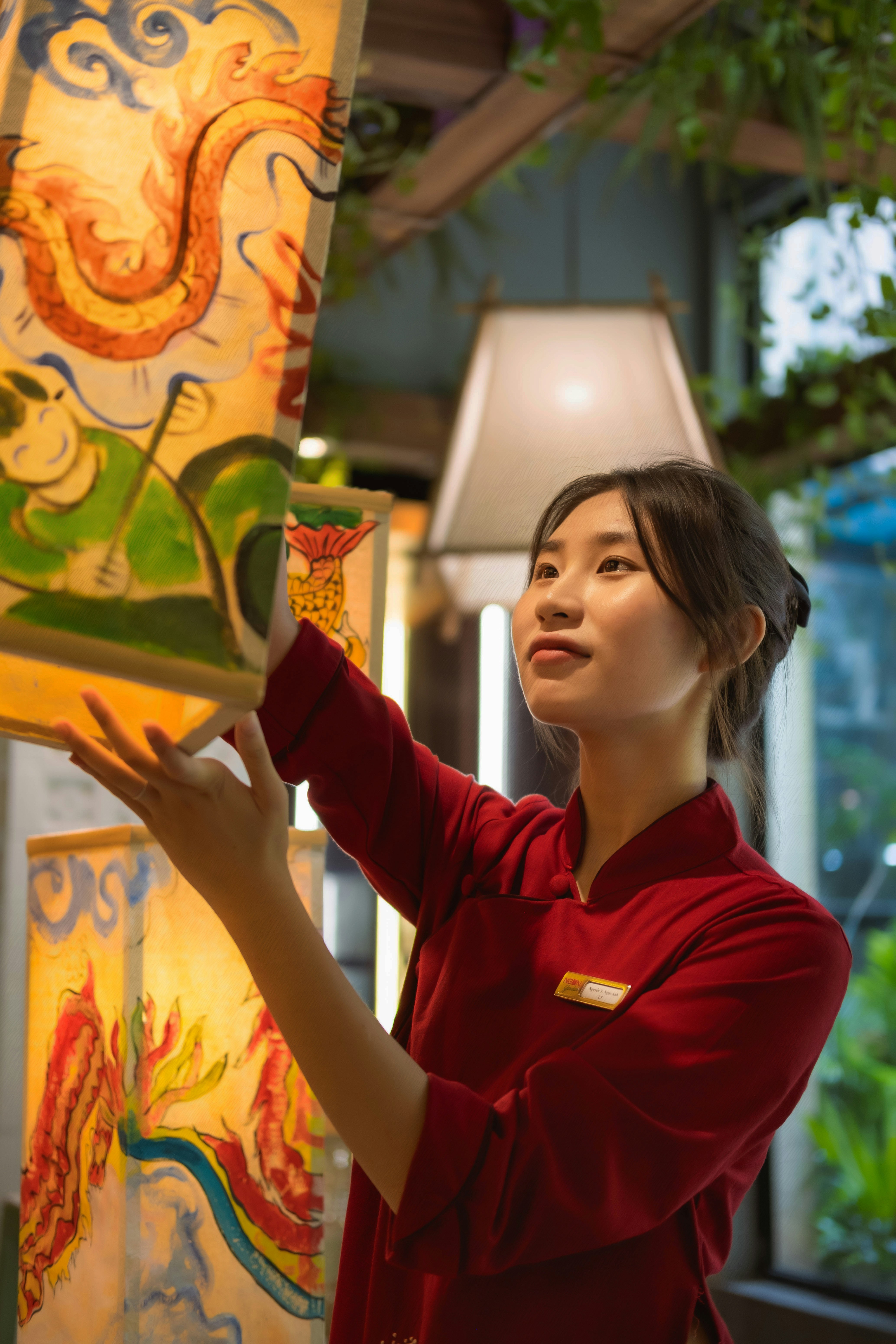 Crimson Attendant Lifts Dragon-Illustrated Lanterns in a Cozy Atelier