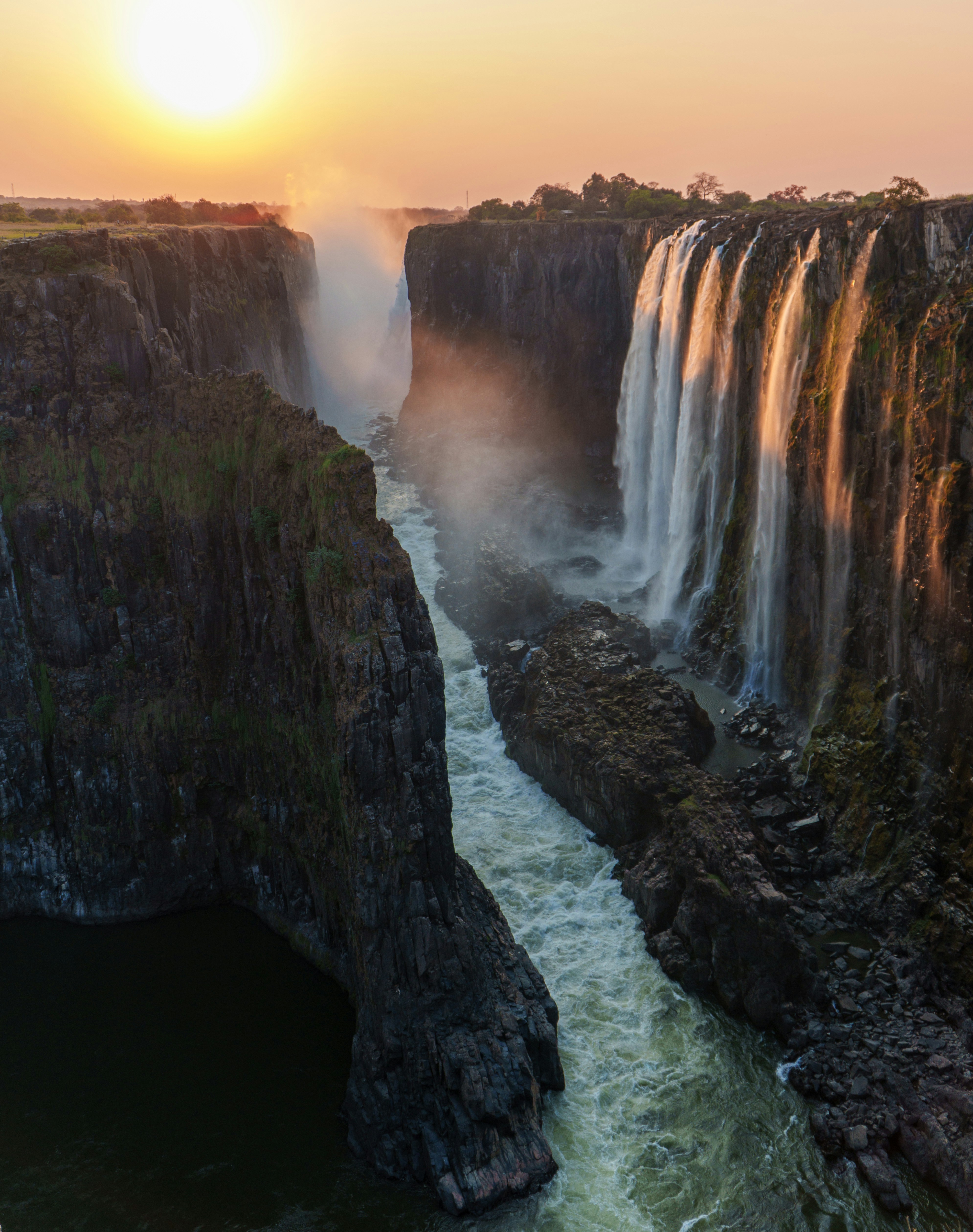 Majestic waterfall cascading into a rocky gorge, illuminated by the warm glow of a setting sun. Mist rises from the water, creating a mystical atmosphere.