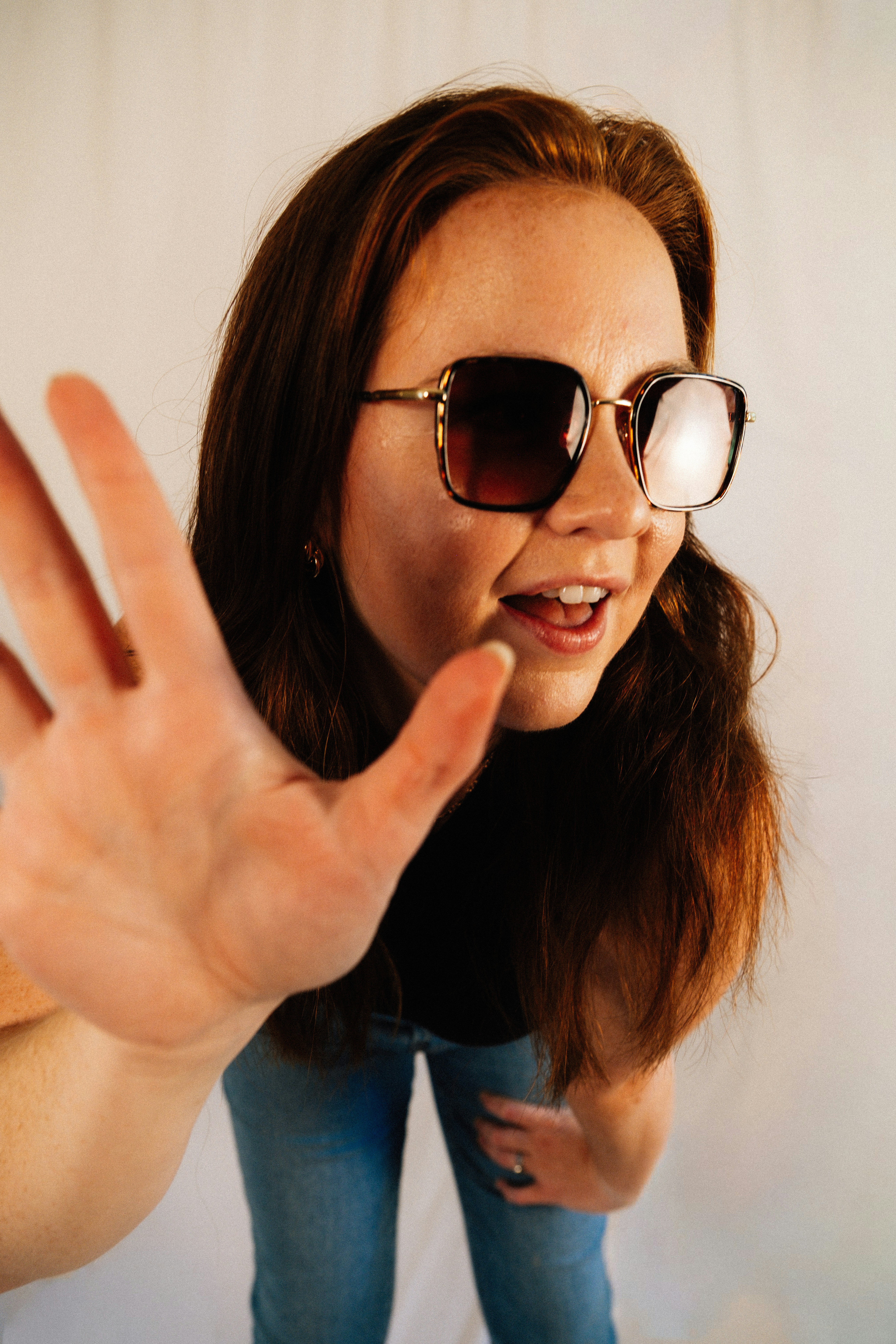 Woman with sunglasses playfully gesturing towards the camera, showcasing a lively expression against a soft backdrop.