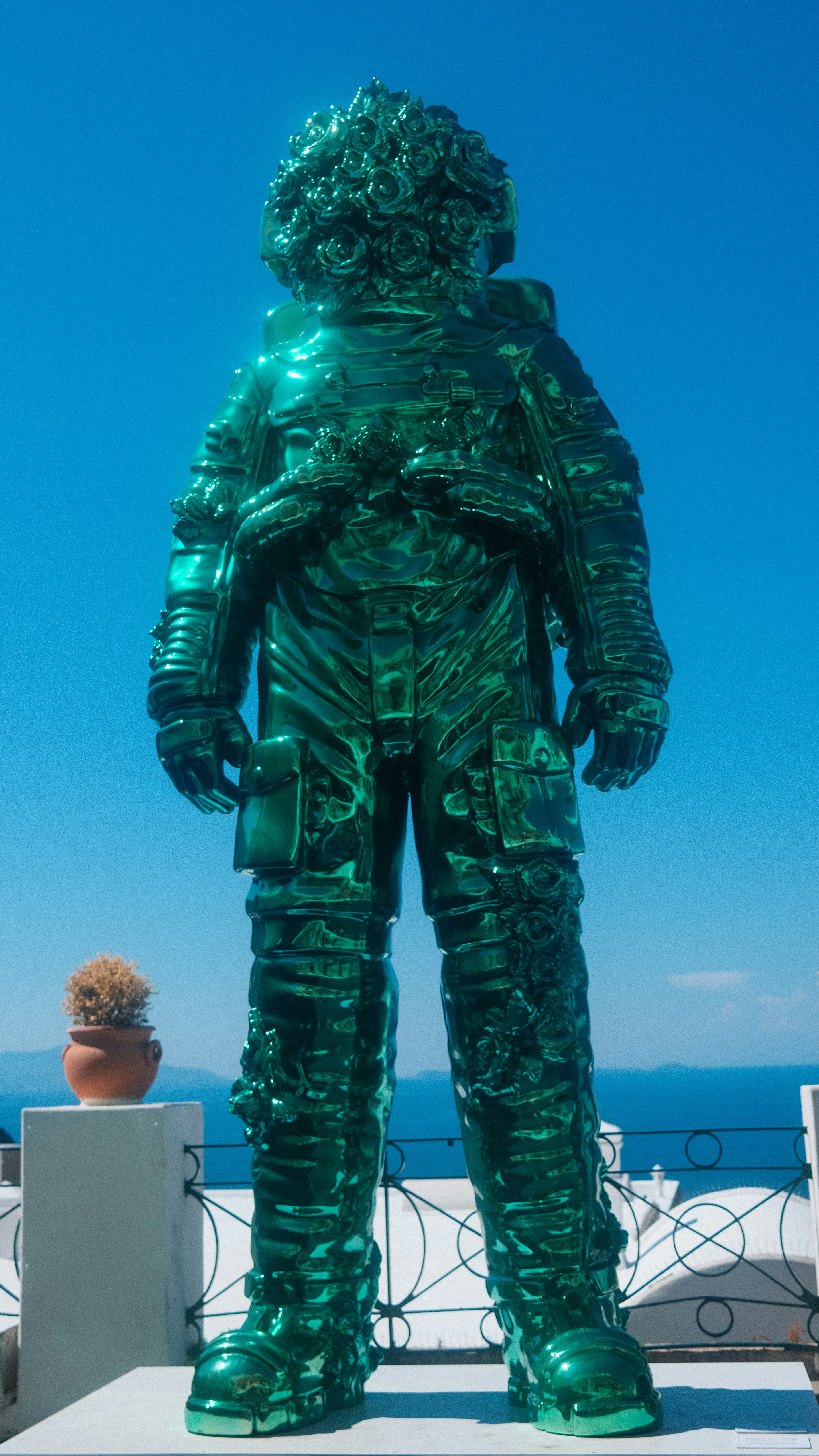Green astronaut sculpture with floral head against blue sky